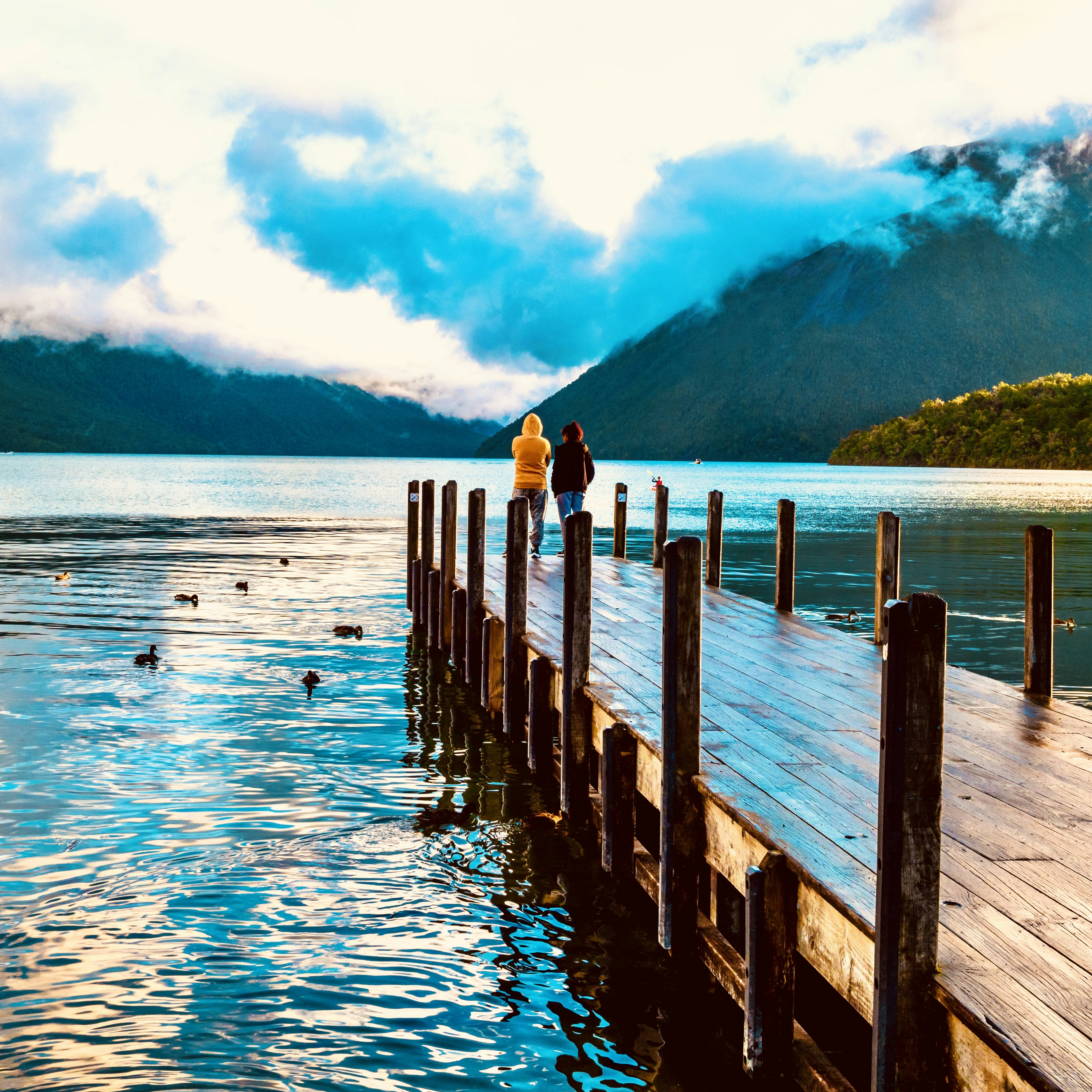 Two figures on a weathered pier reach toward a turquoise lake framed by misty mountains