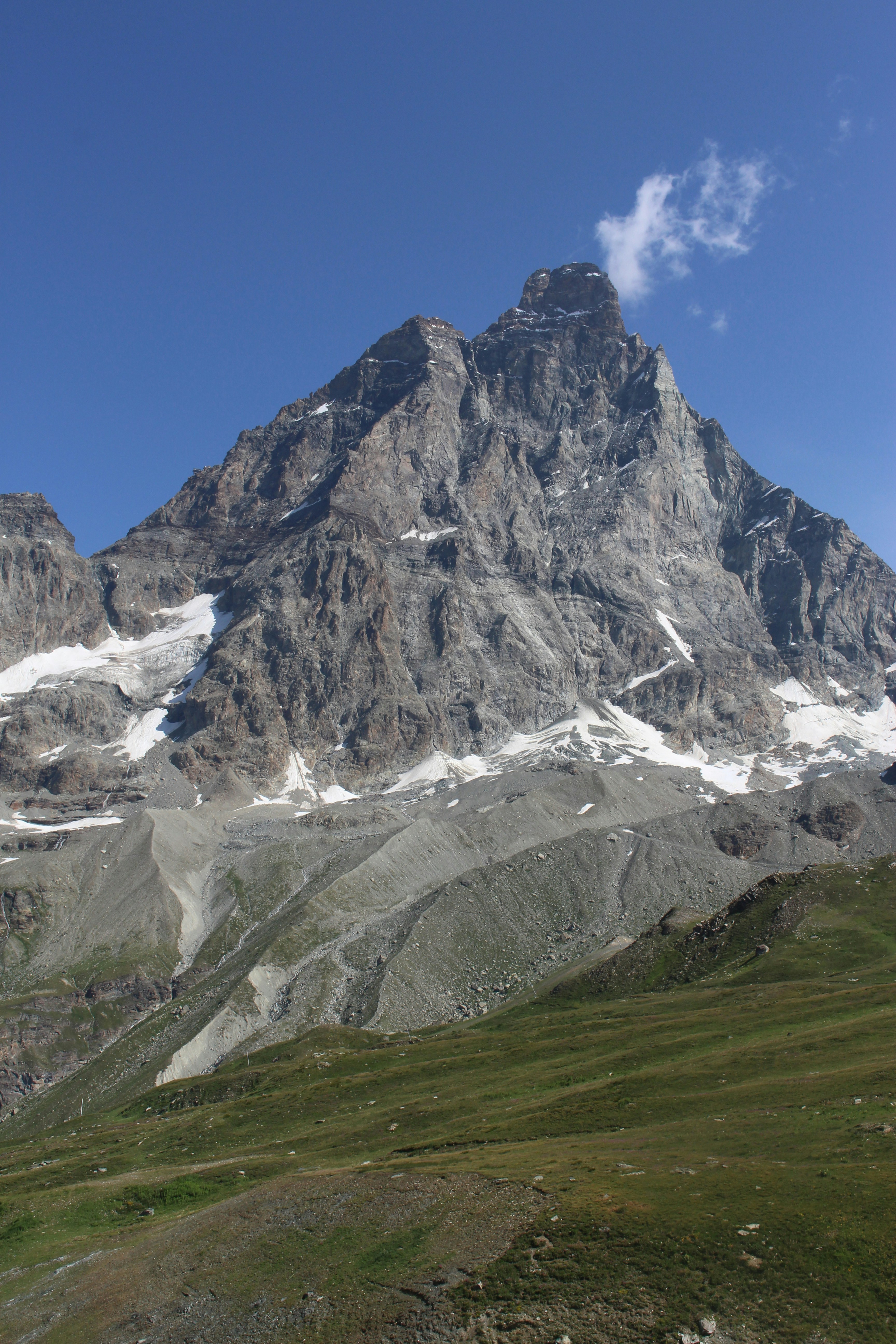 Jagged mountain summit with patches of snow rises against a deep blue sky; the lower slopes flow into green meadow and rocky terrain.