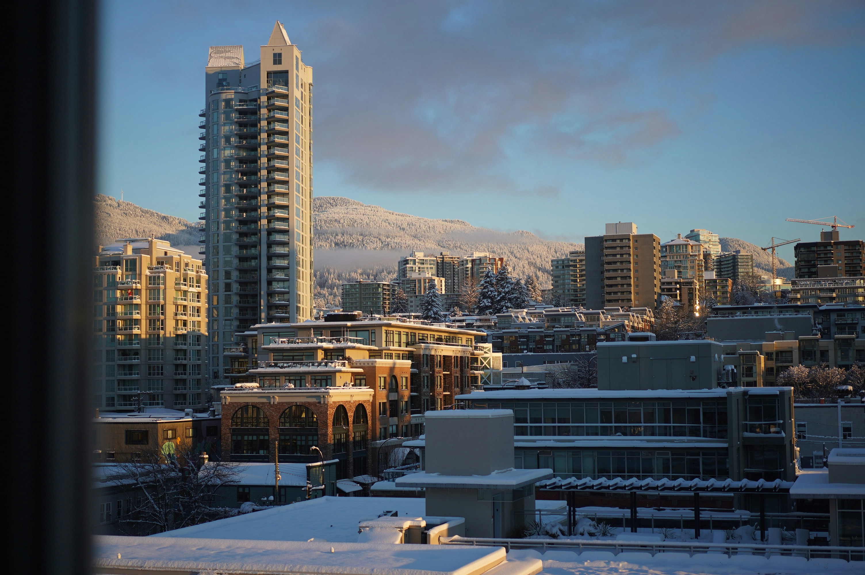Snow-coated cityscape bathed in warm late-afternoon light, with mid-rise and high-rise buildings clustered at the horizon and a snow-blanketed foreground.