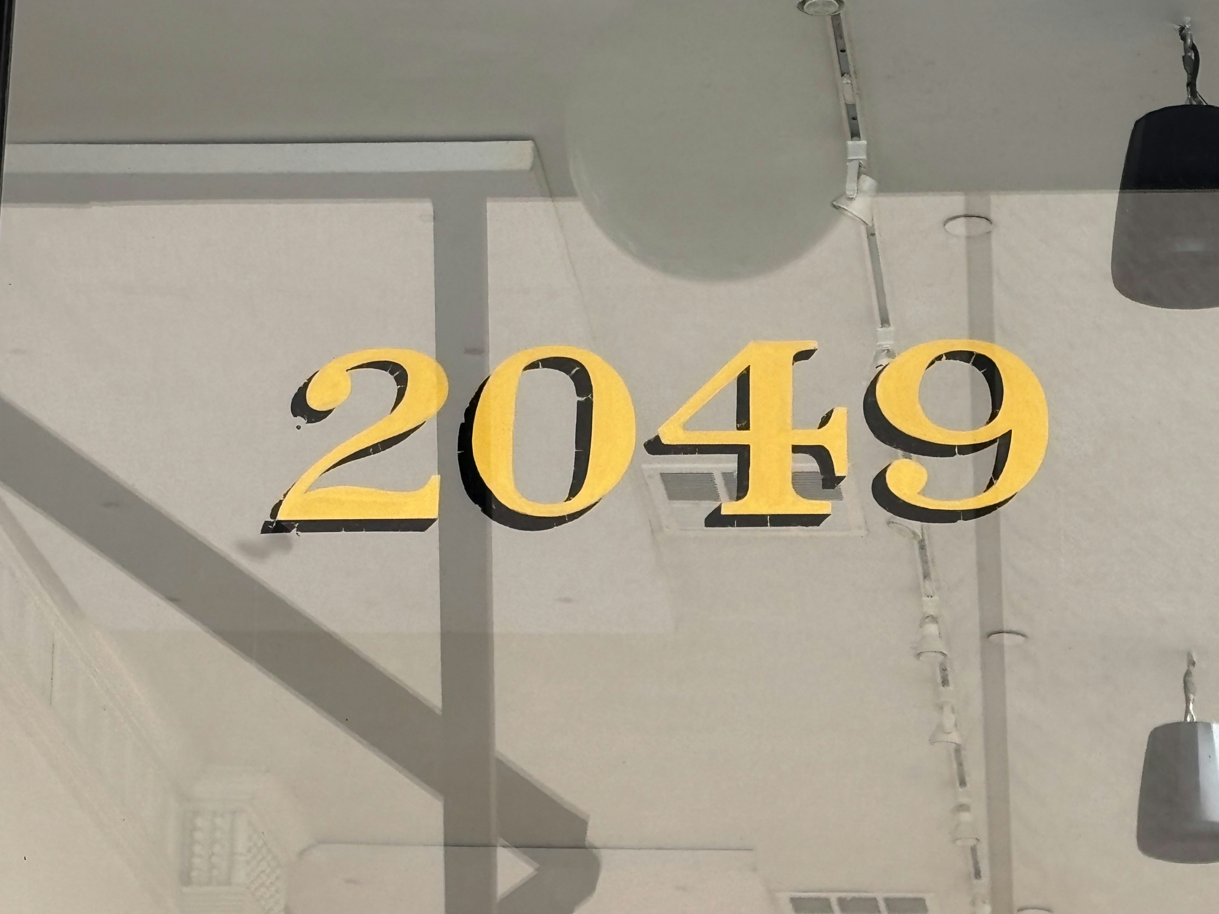 Close-up of bold golden digits '2049' mounted on a pale wall with soft architectural shadows and industrial fixtures in the background.