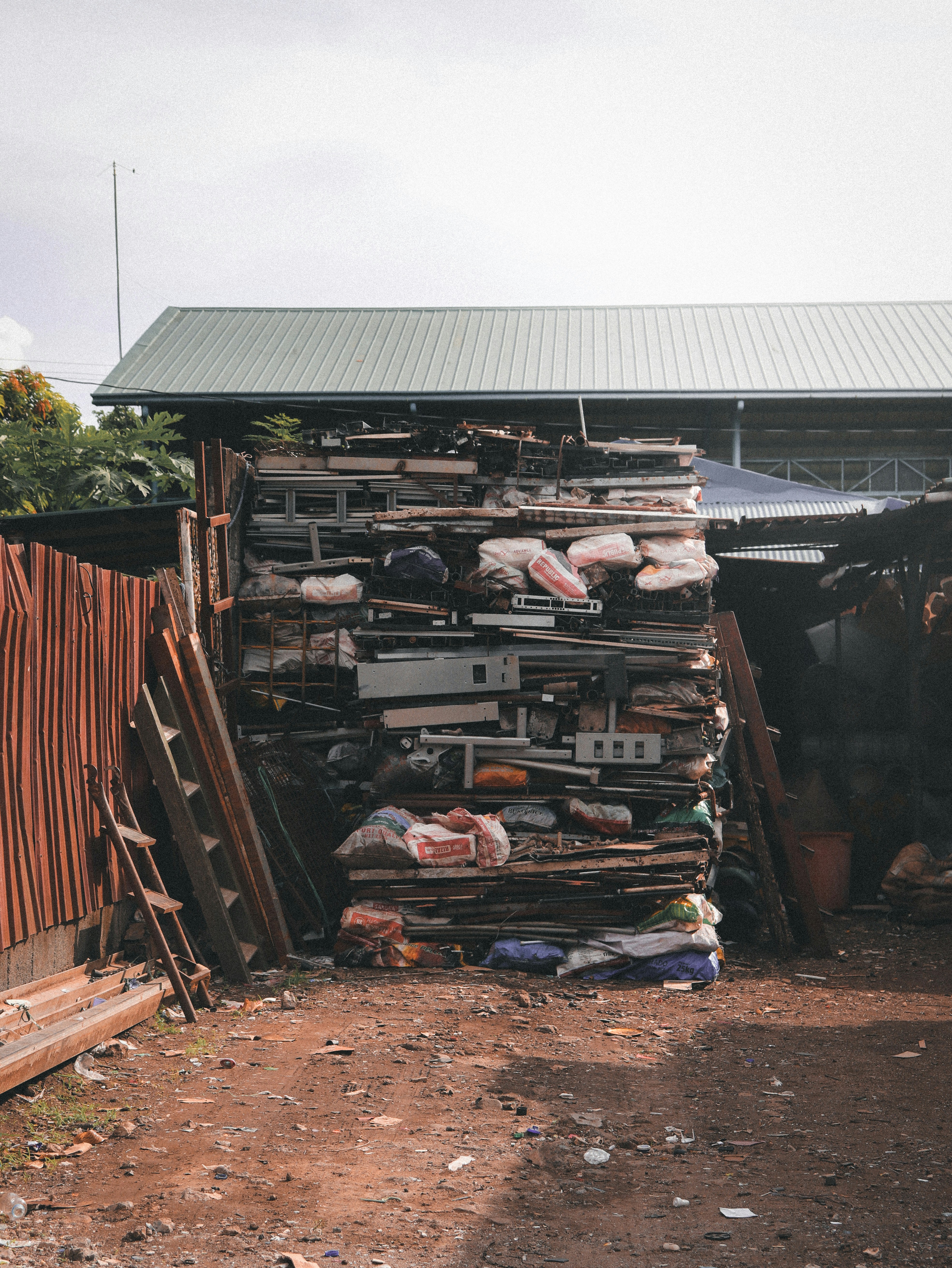 Heap of discarded materials and pallets stacked under a corrugated roof, with a dirt ground and scattered debris in the foreground.