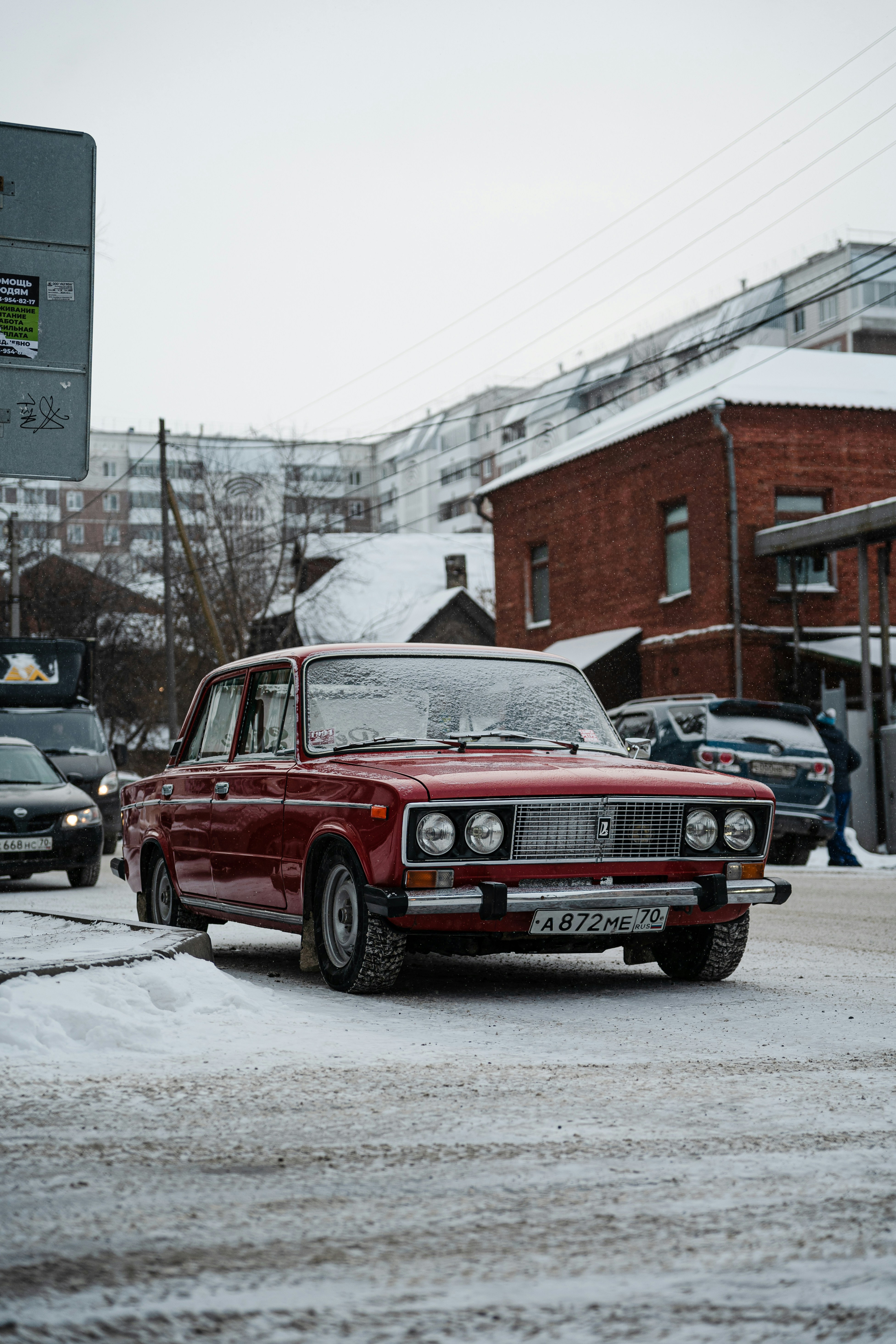 A classic red vintage sedan is parked on a snowy city street during winter. The car stands out against the muted surroundings, with snow-covered roads, brick buildings, apartment blocks, and overcast skies creating a cold urban atmosphere. Other vehicles and utility poles in the background add to the everyday street scene, while the vintage car becomes the clear focal point.