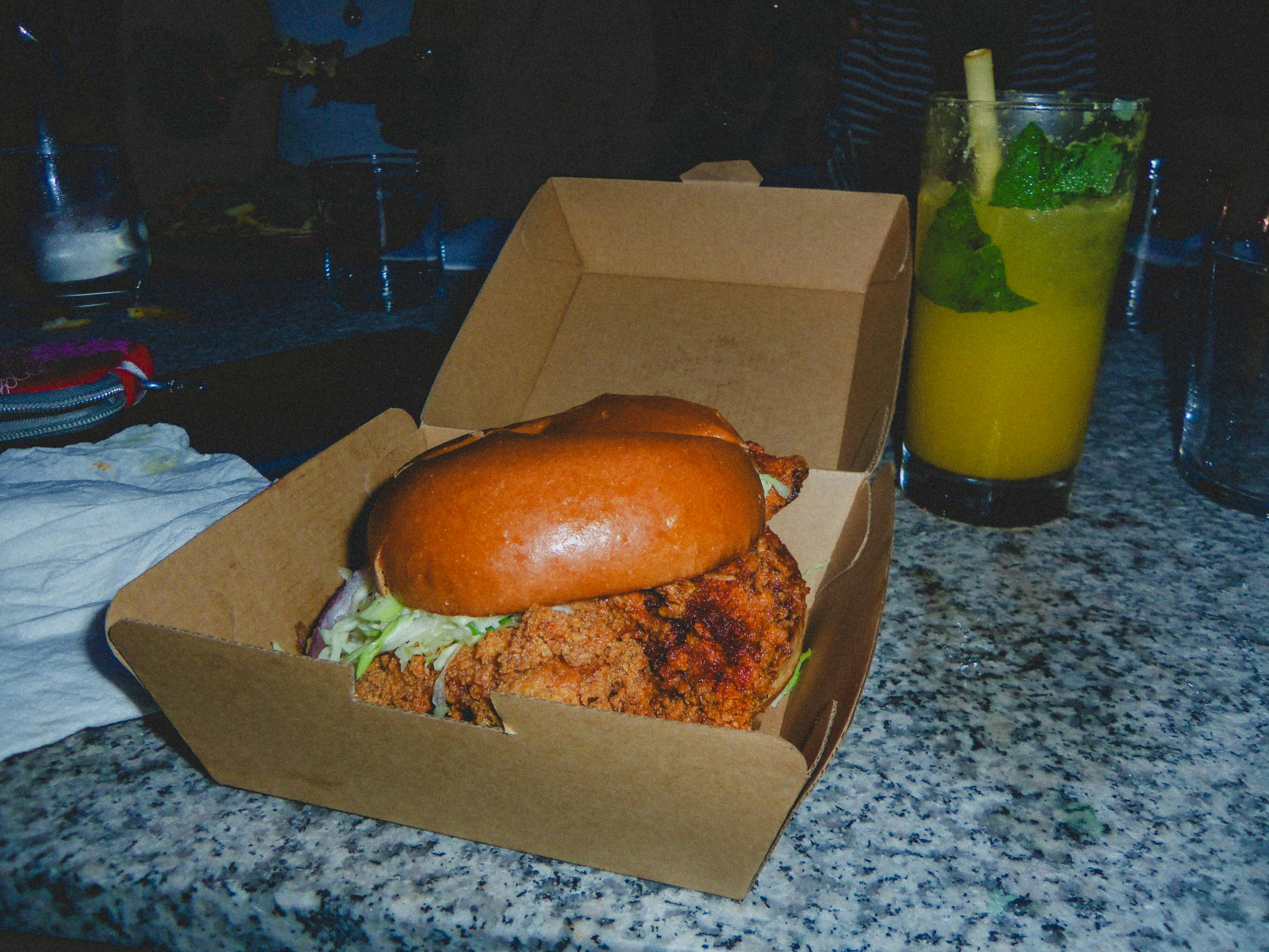Crispy chicken sandwich in a brown box sits on a granite table beside a tall yellow beverage with mint; casual dining scene with dim lighting.