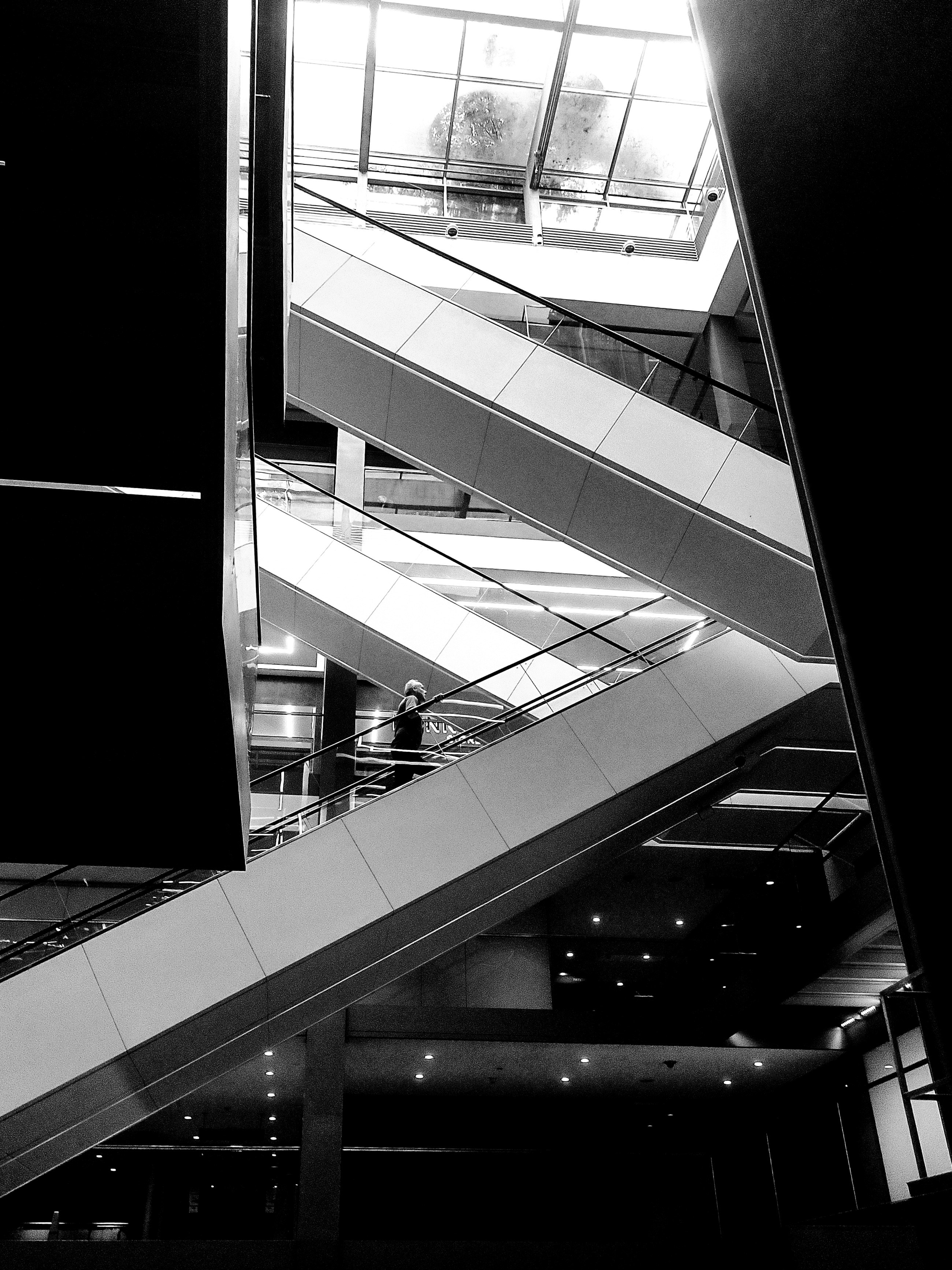 A high-contrast black and white architectural photograph showing a lone silhouette riding an intricate system of crisscrossing escalators inside a modern building. The dramatic shadows and sharp diagonal lines create a powerful, graphic composition.