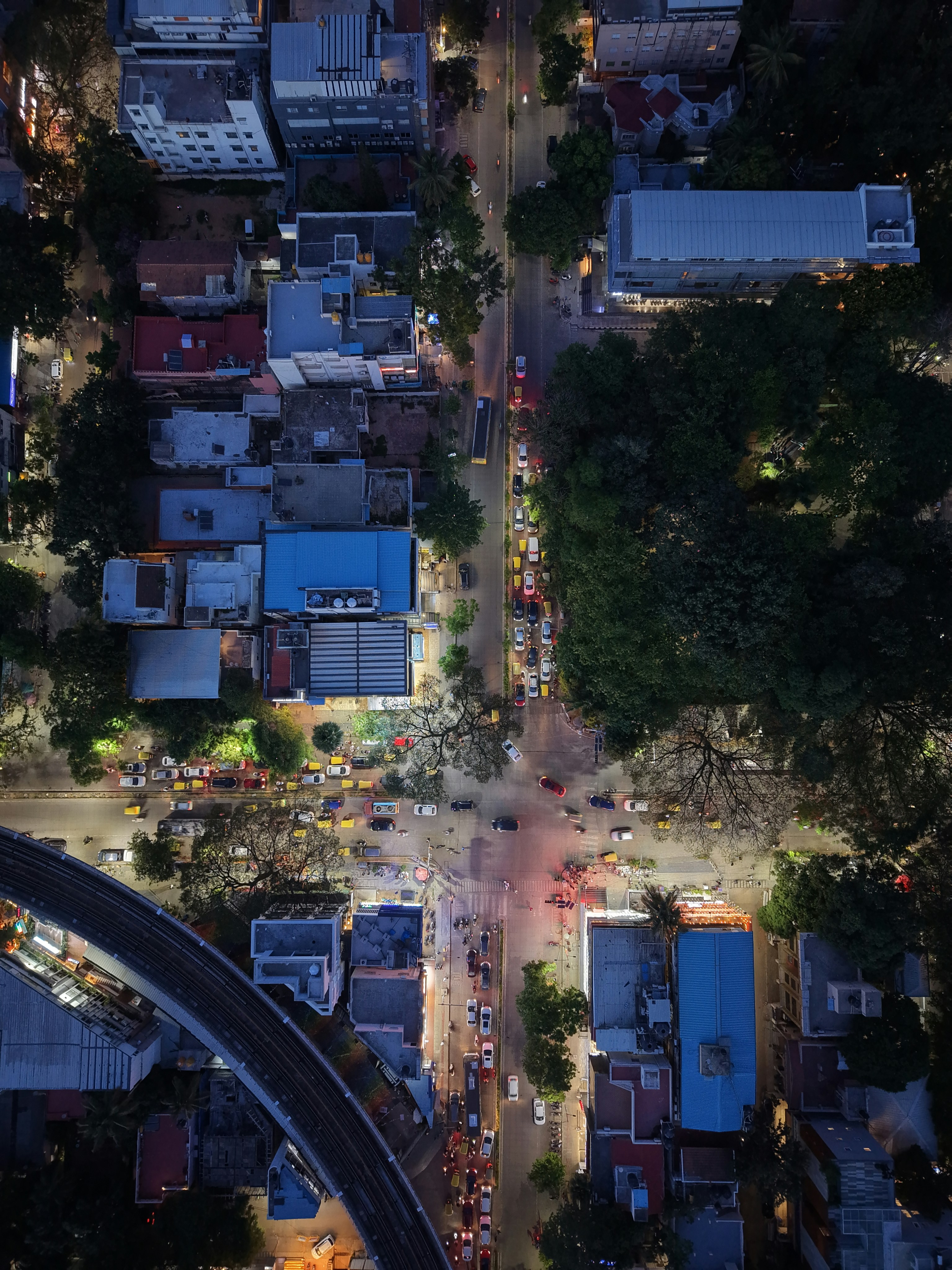 Aerial view of a city street intersection lined with colorful buildings and vehicles, captured in twilight with dense tree canopy on one side and a curved elevated structure on the left.