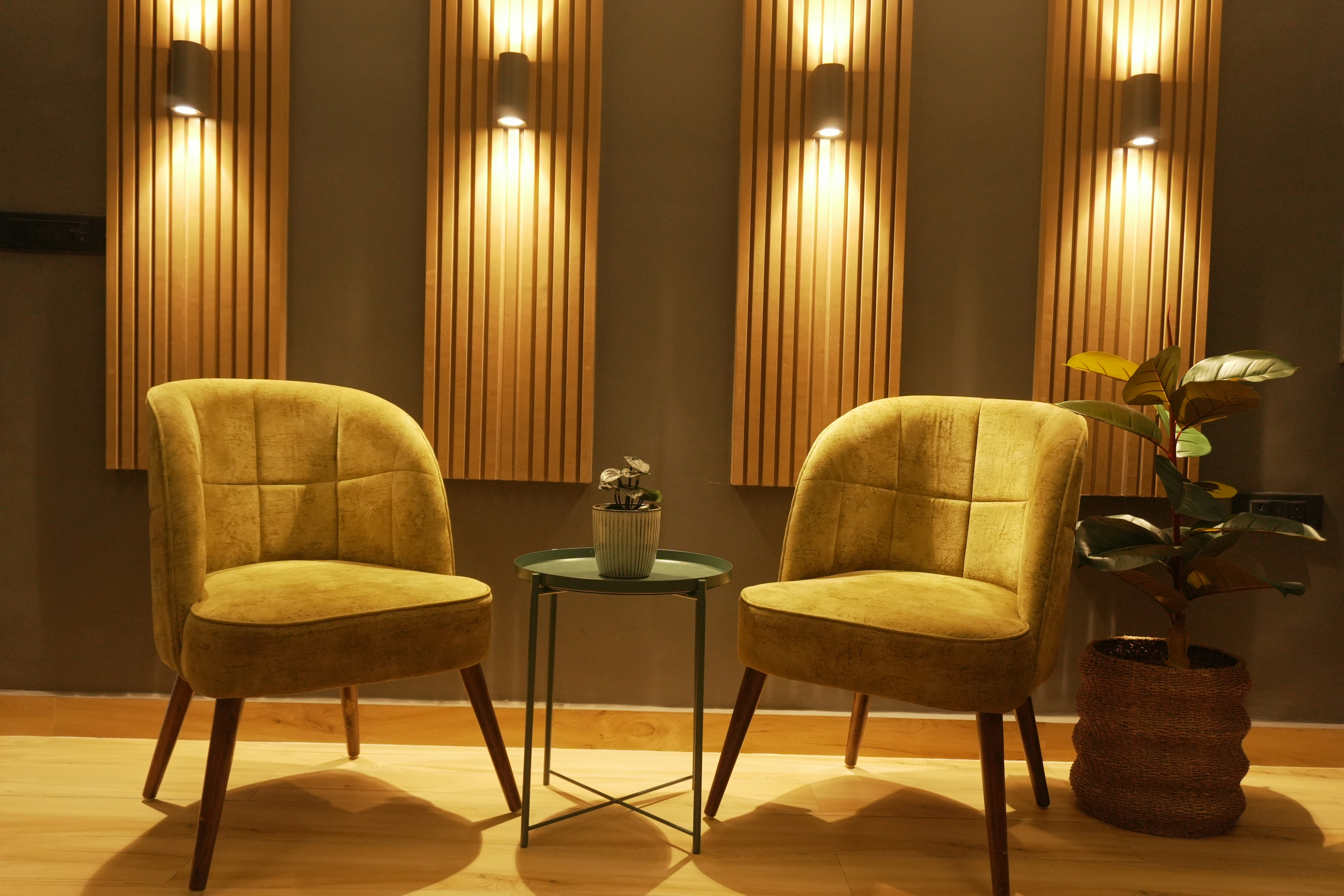 Two mustard-yellow lounge chairs flank a small round table under warm wall-mounted lighting in a minimal studio setup.