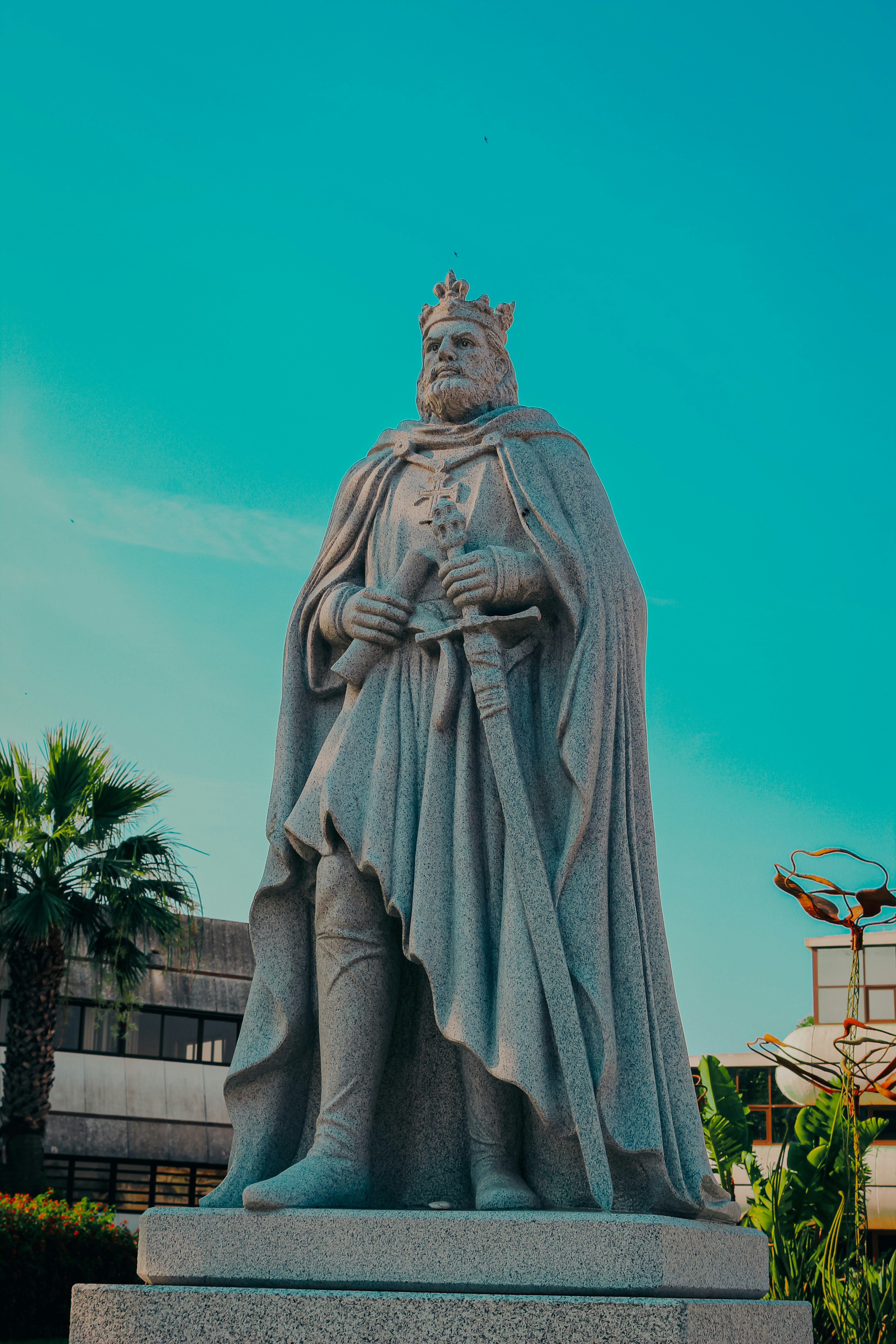 Granite statue of a crowned king holding scepter, standing outdoors with palm trees and modern buildings in the background, captured with clear blue sky.