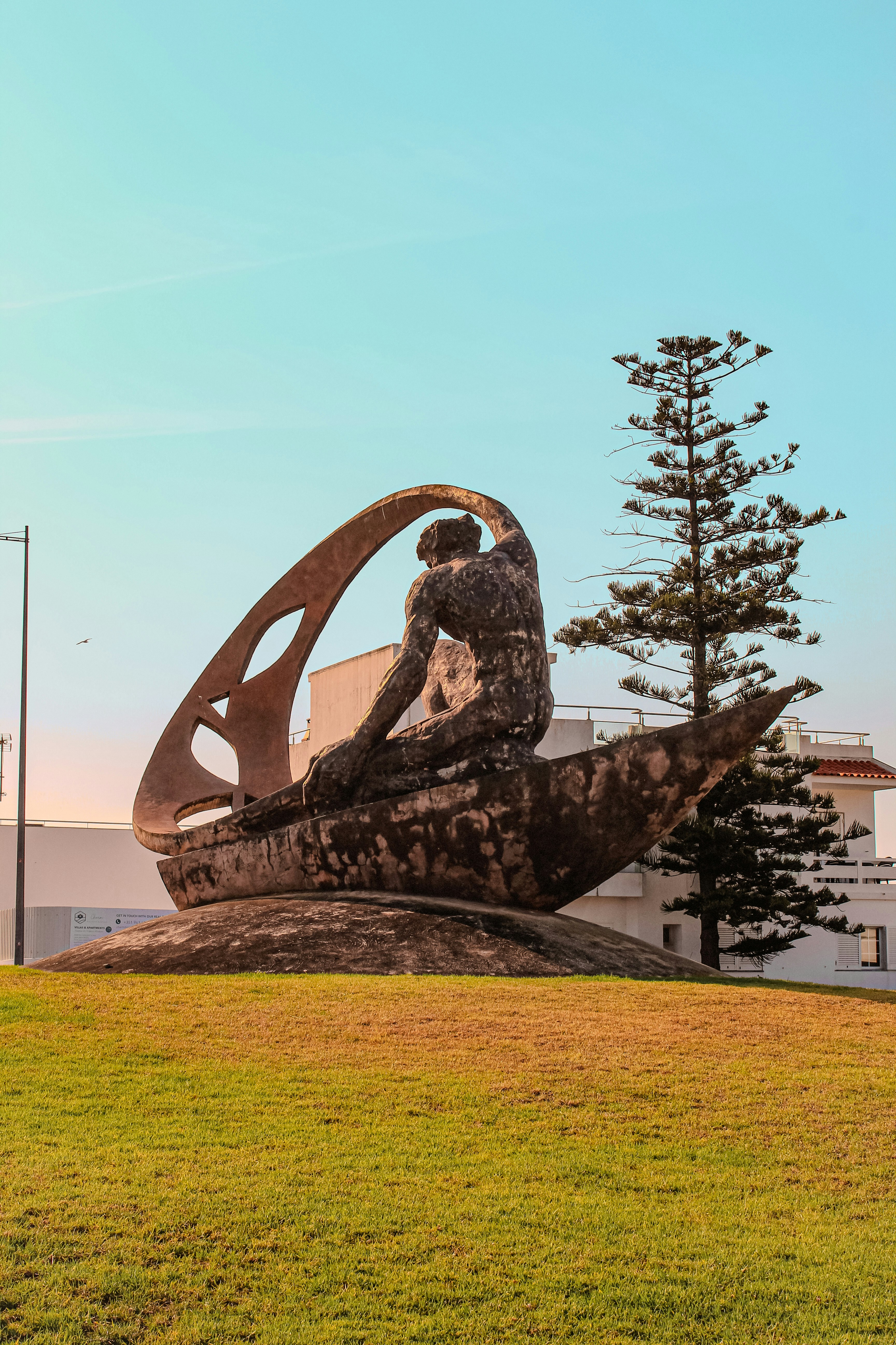 Rusty sculpture of a seated figure inside a curved hull, set on a grassy mound with a modern building and sparse tree in the background; a quiet coastal plaza scene.