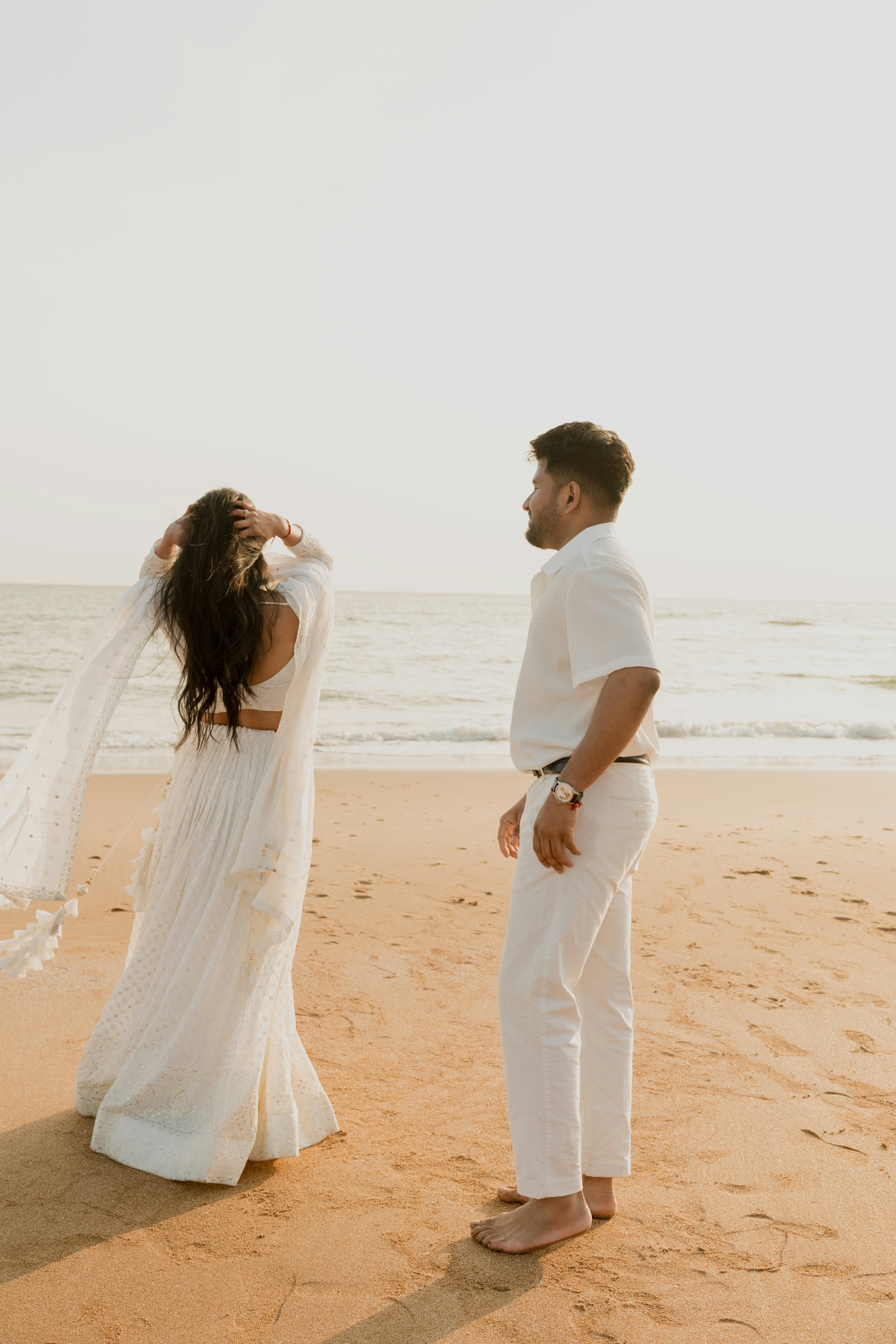 A couple stands on a sunlit beach, the woman adjusting her light, flowing dress while the man faces her near the shoreline; the scene emphasizes calm, warm tones and a minimalist horizon. It is a candid lifestyle photograph.