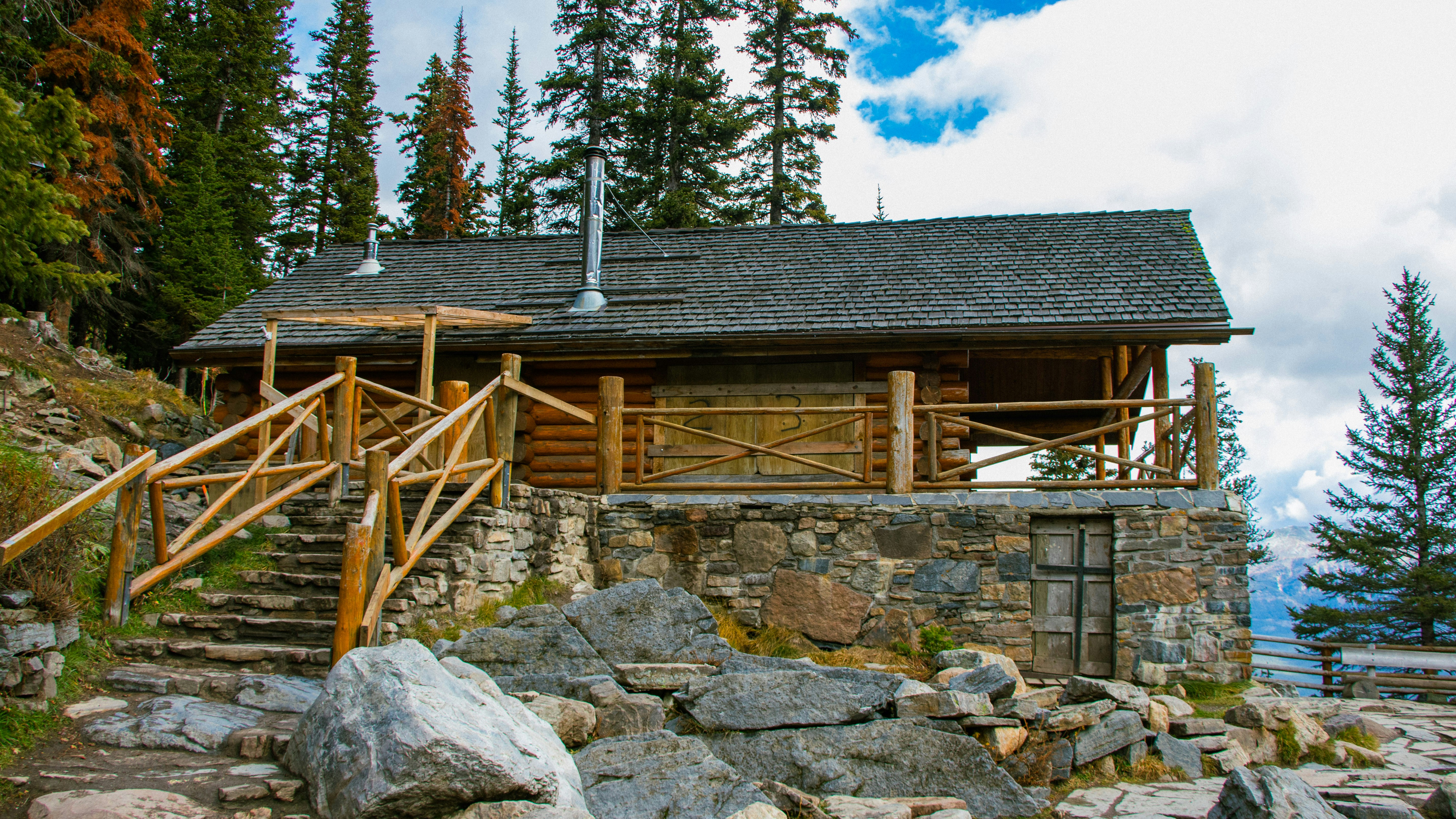 A rustic log cabin perched on a stone terrace beside a rocky staircase amid evergreen pines. The scene emphasizes the rugged mountain shelter set against a bright sky.