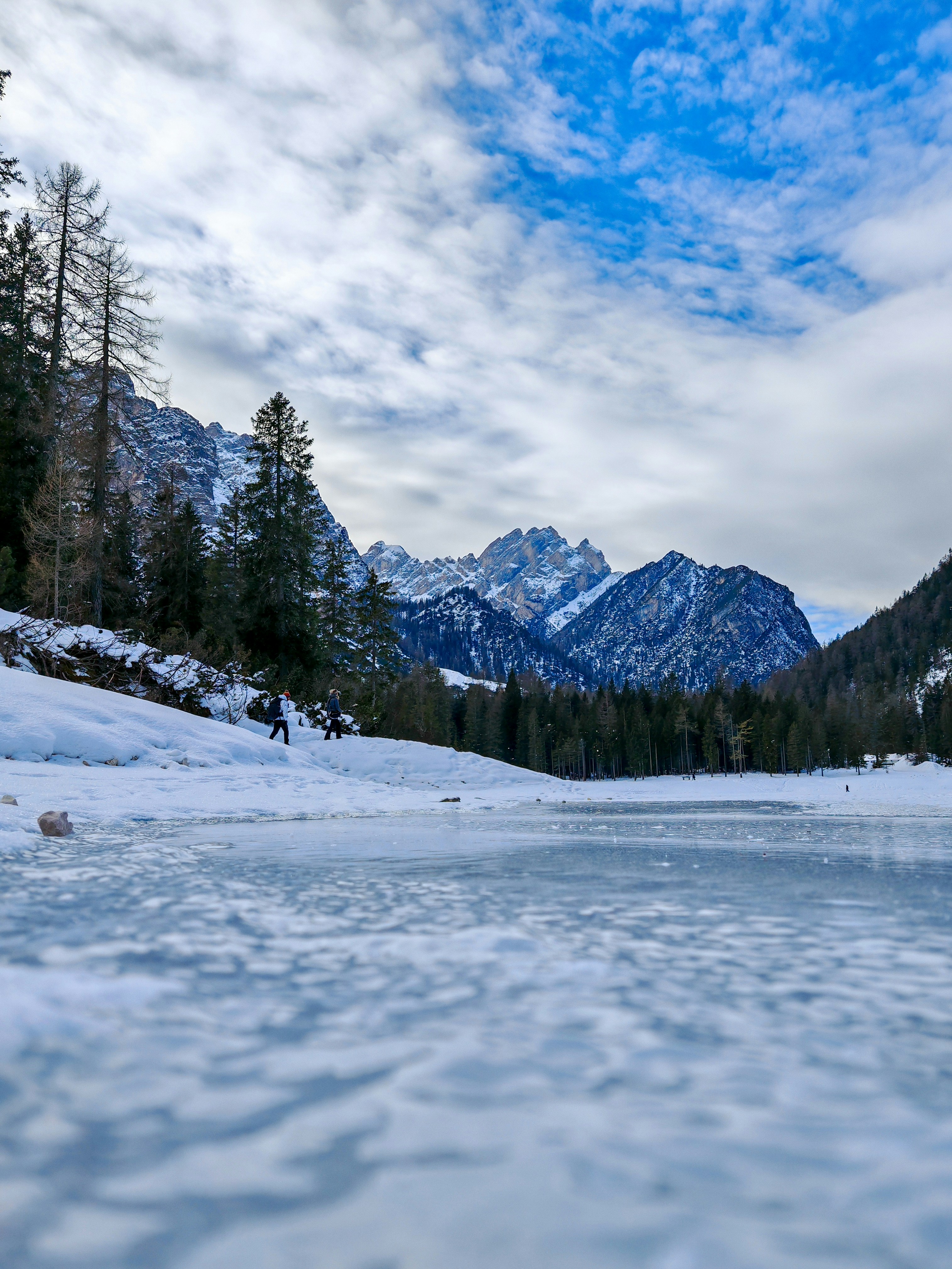 Snow-covered lake with distant hikers along the shore, pine trees lining the left, and jagged alpine peaks under a partly cloudy sky.