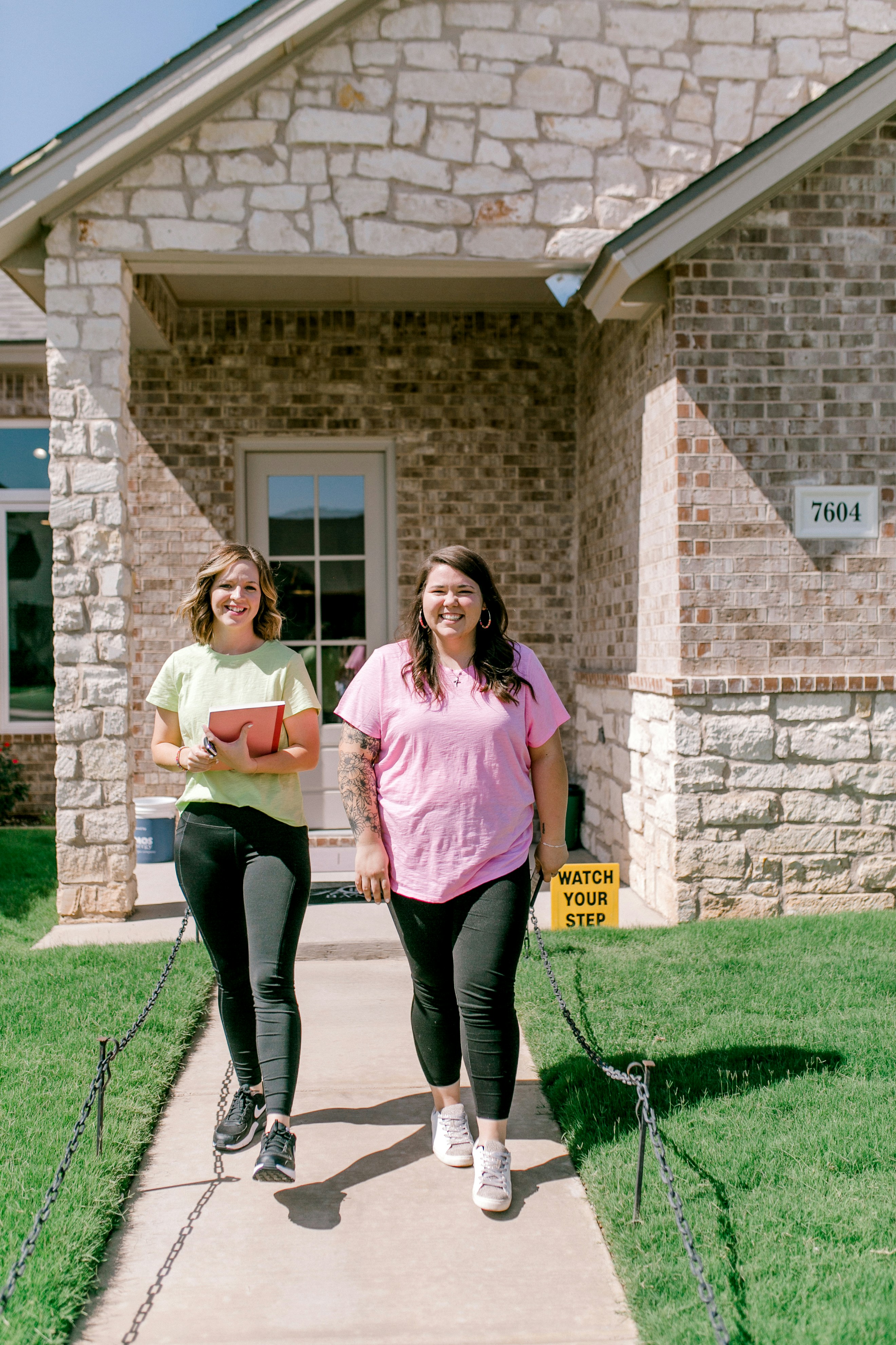 Two smiling women walk on a path from a house.