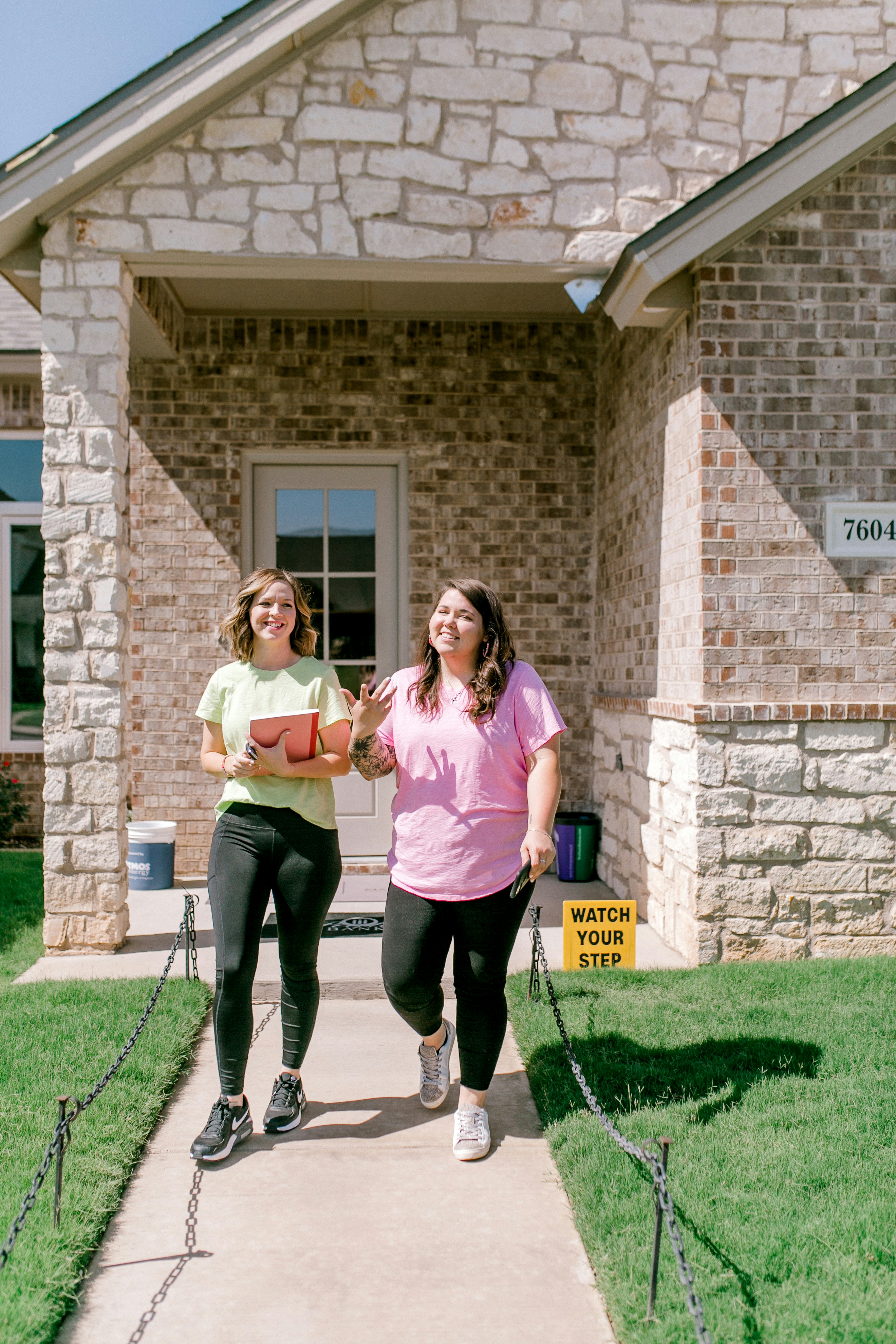 Two friends leave a sunlit campus building, laughter spilling onto the sidewalk