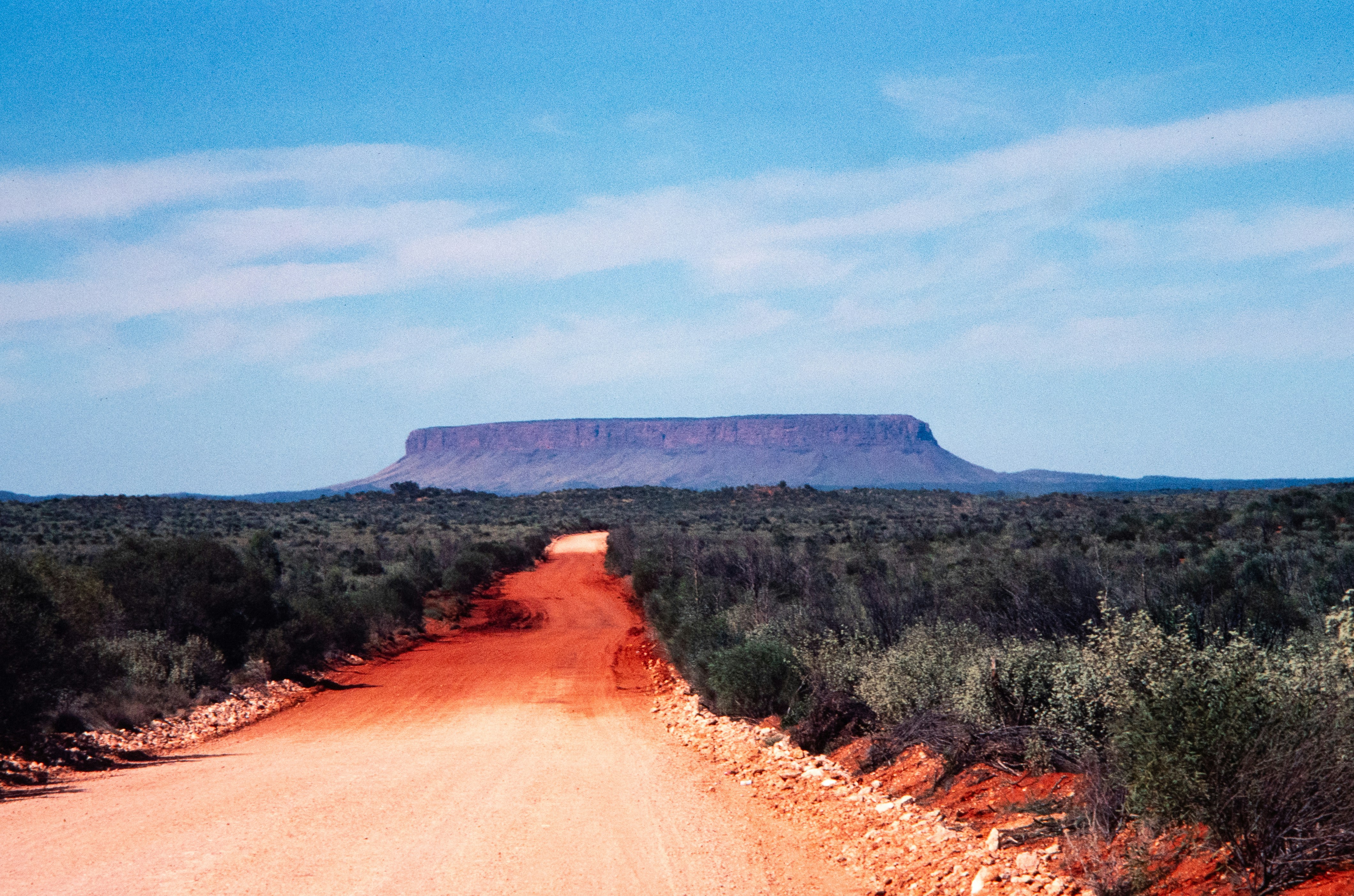 Dusty red road through scrubland towards a distant mesa.