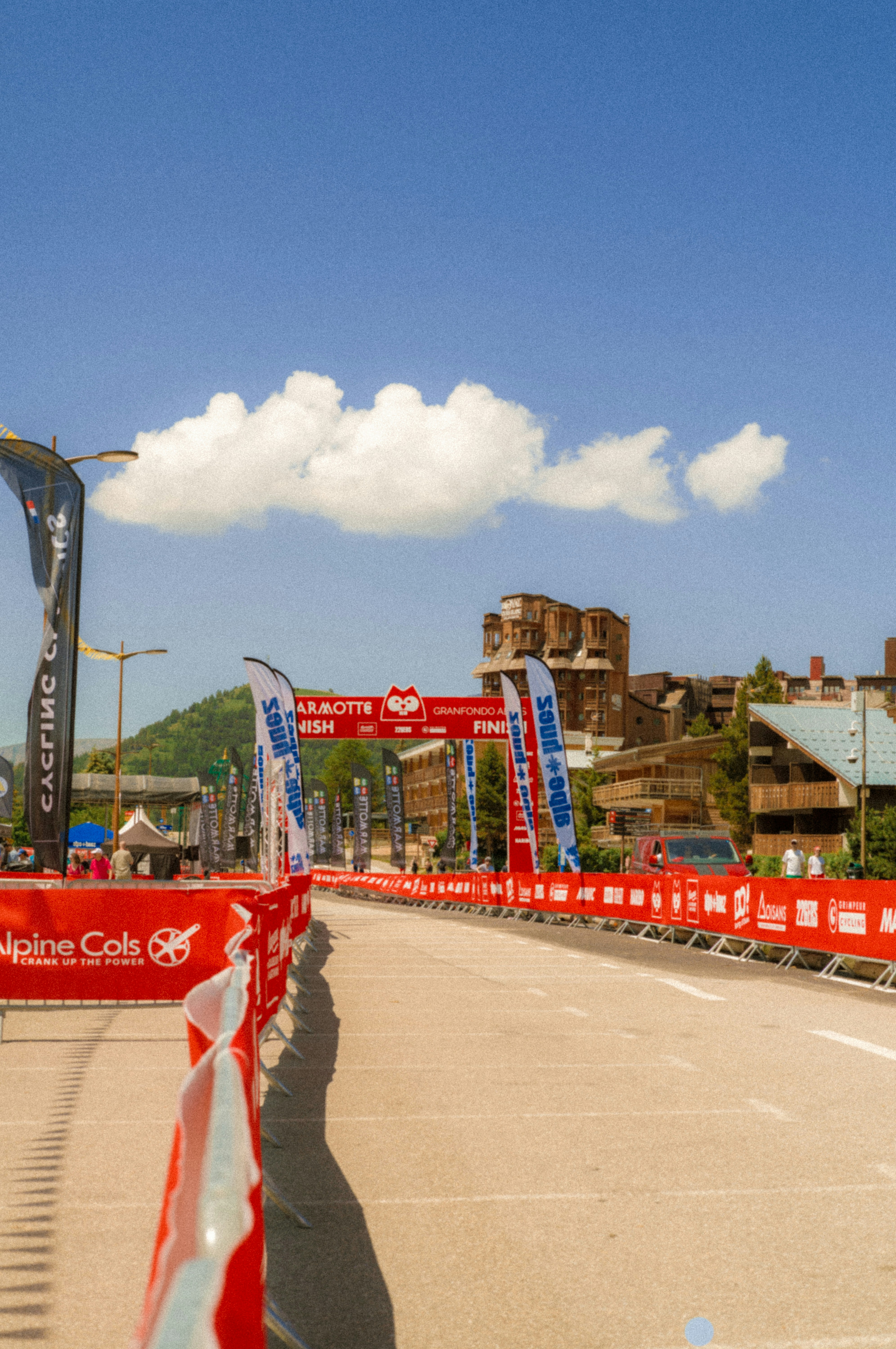 Racecourse lane lined with red barriers and sponsor banners leading toward a distant finish arch against a bright blue sky.