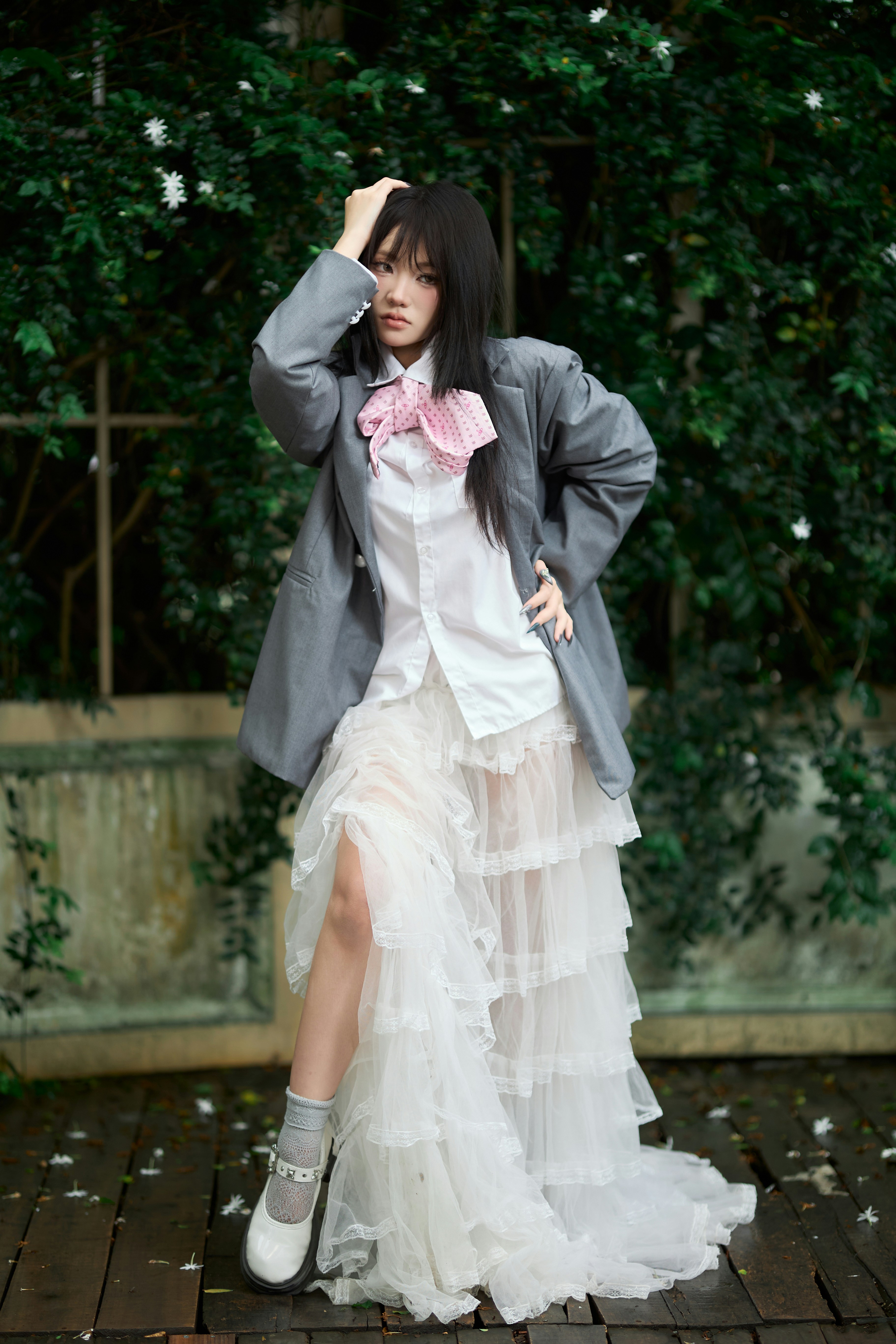 Young woman in a layered white dress and gray jacket poses amid green foliage; soft natural light emphasizes texture and movement of the fabric.