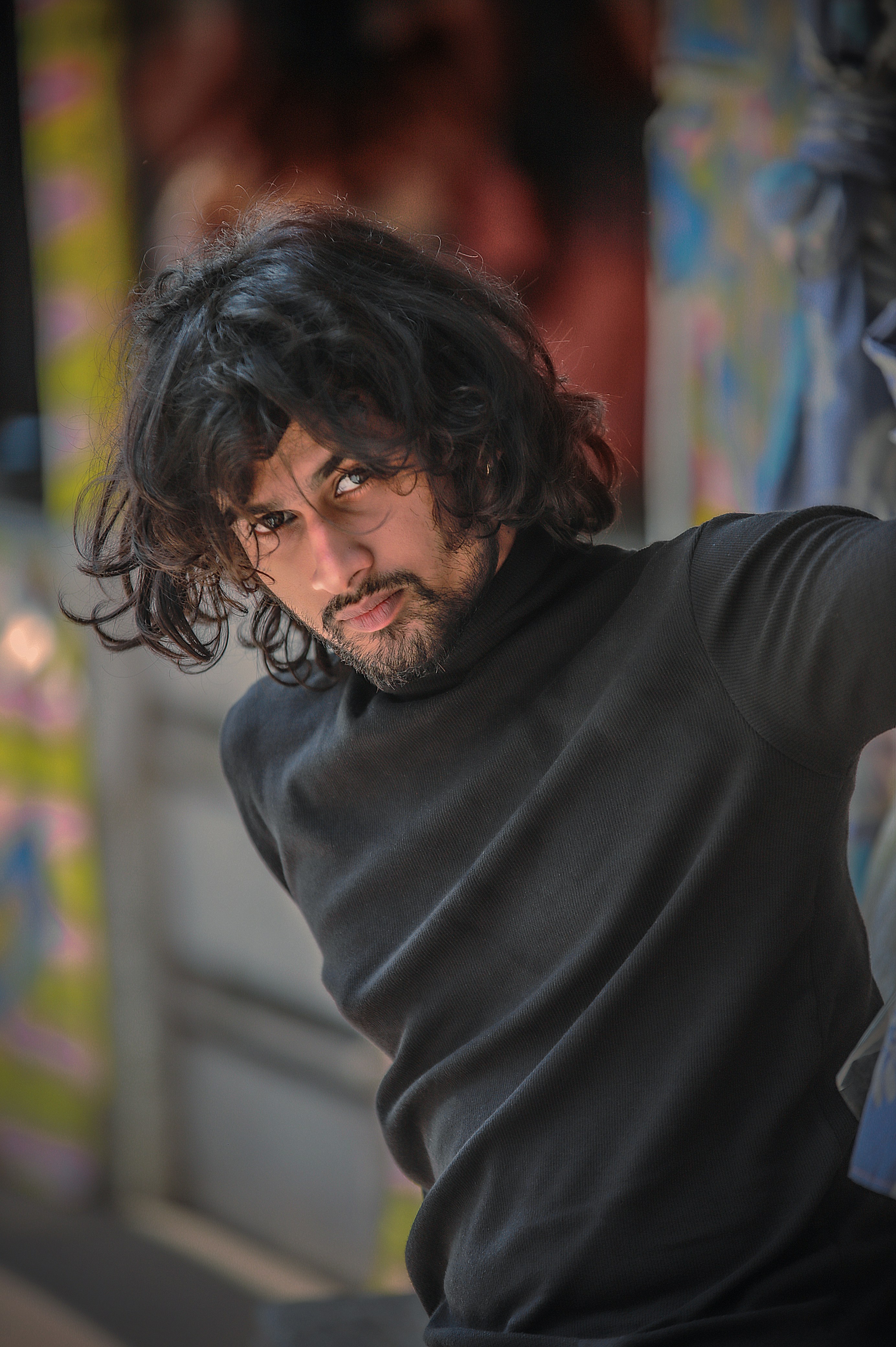 Close-up portrait of a man with tousled dark hair and a trimmed beard, wearing a black turtleneck, leaning against a colorful urban backdrop. The focus centers on his intense gaze and relaxed pose.