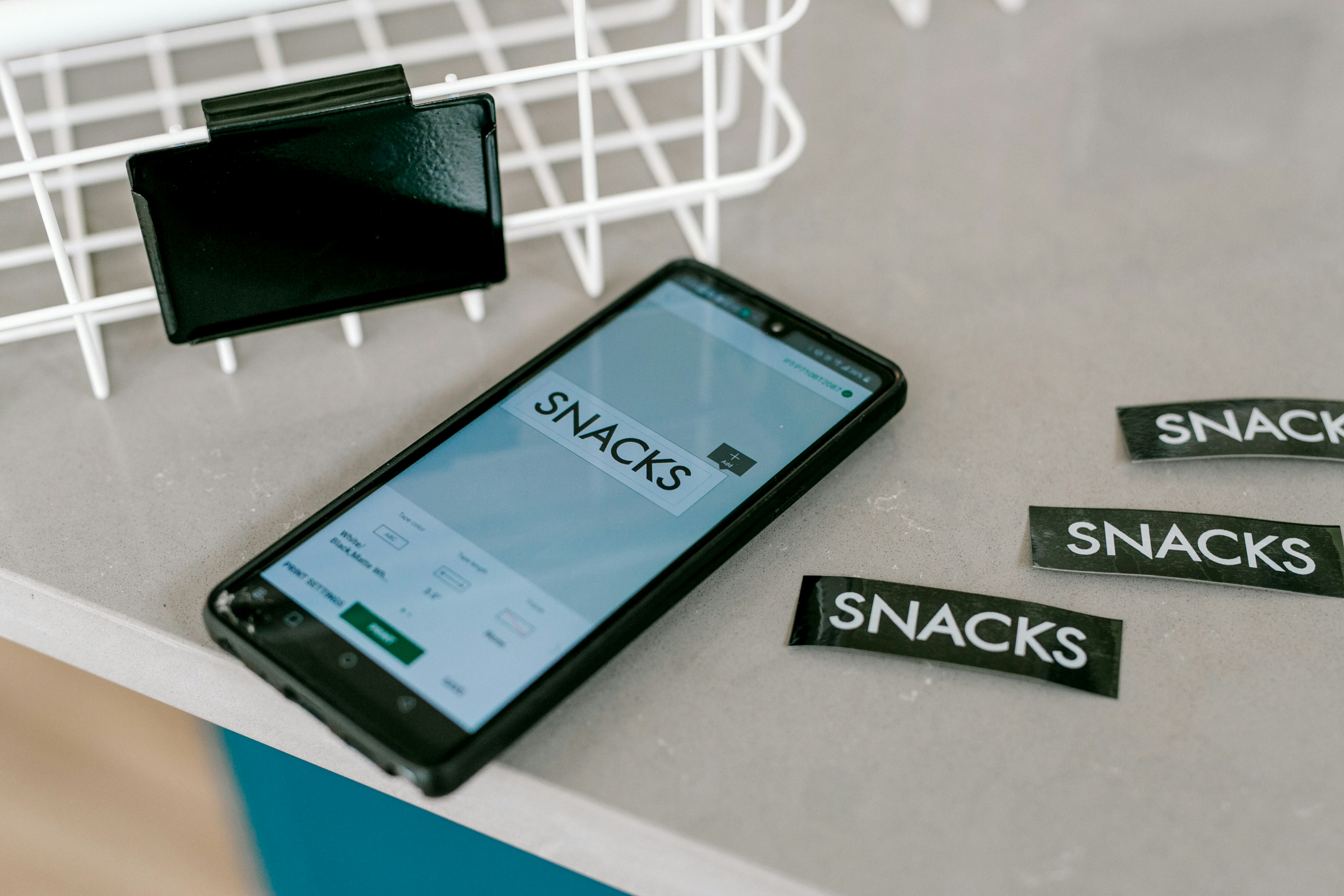 Smartphone screen shows SNACKS app on a countertop with snack labels nearby; a small white rack and wallet are in the background, suggesting a cafe or store setting.