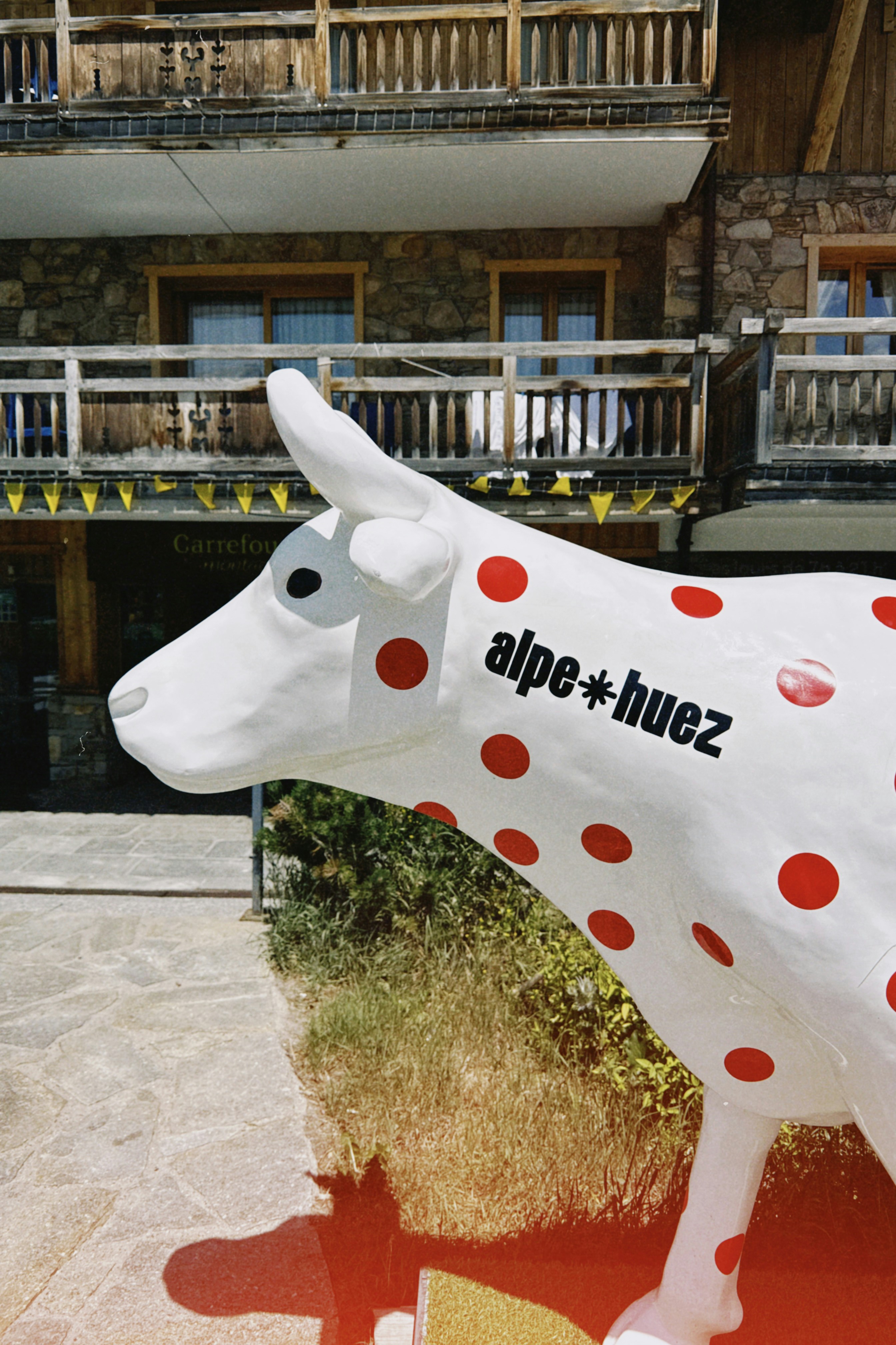 White cow sculpture with red polka dots and 'alpe*huez' lettering stands in front of a stone chalet with wooden balconies, under a sunlit pavement and sparse greenery.