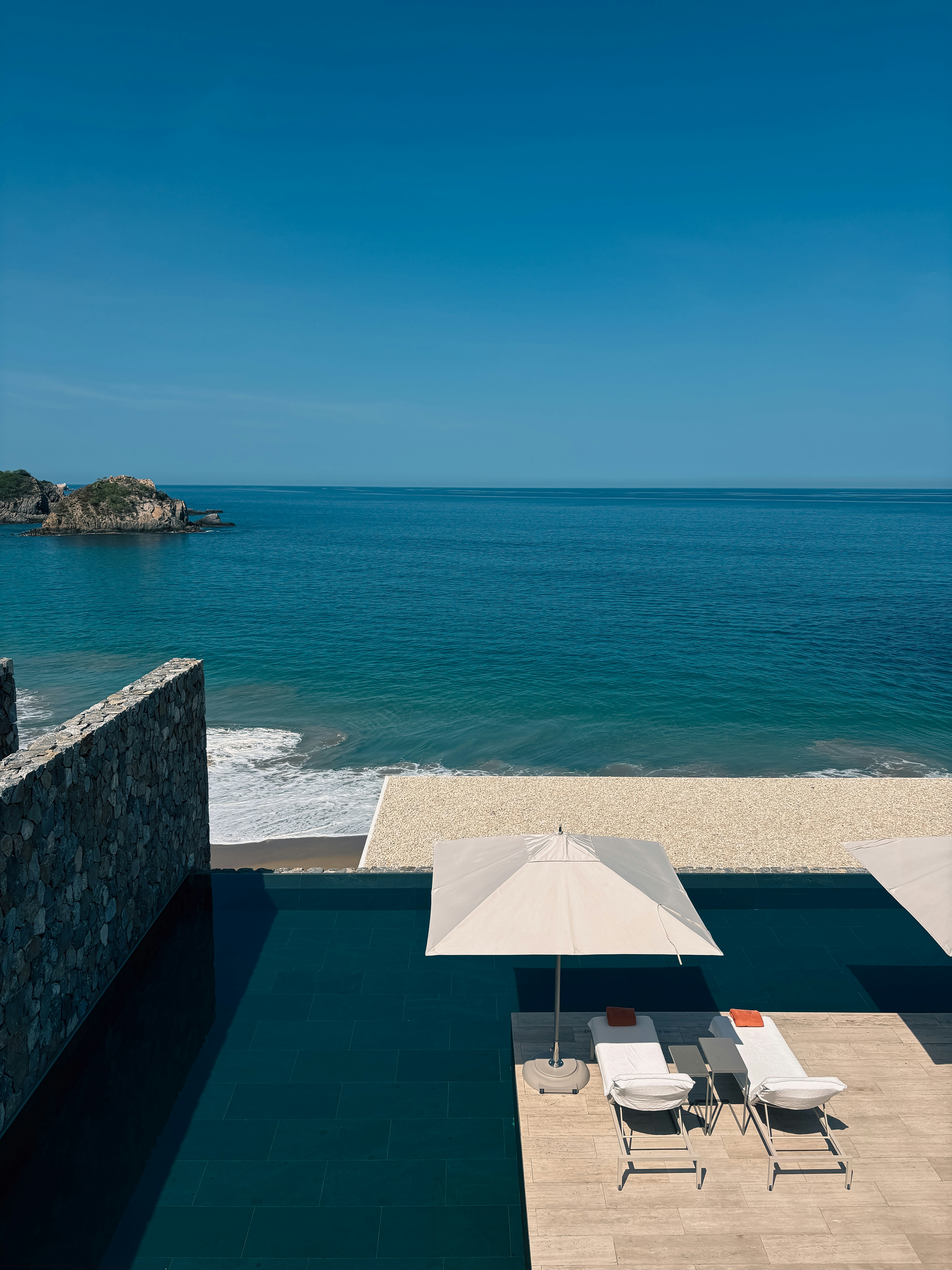 An infinity pool with an umbrella and loungers overlooks the Pacific Ocean and a rocky islet in Mexico.