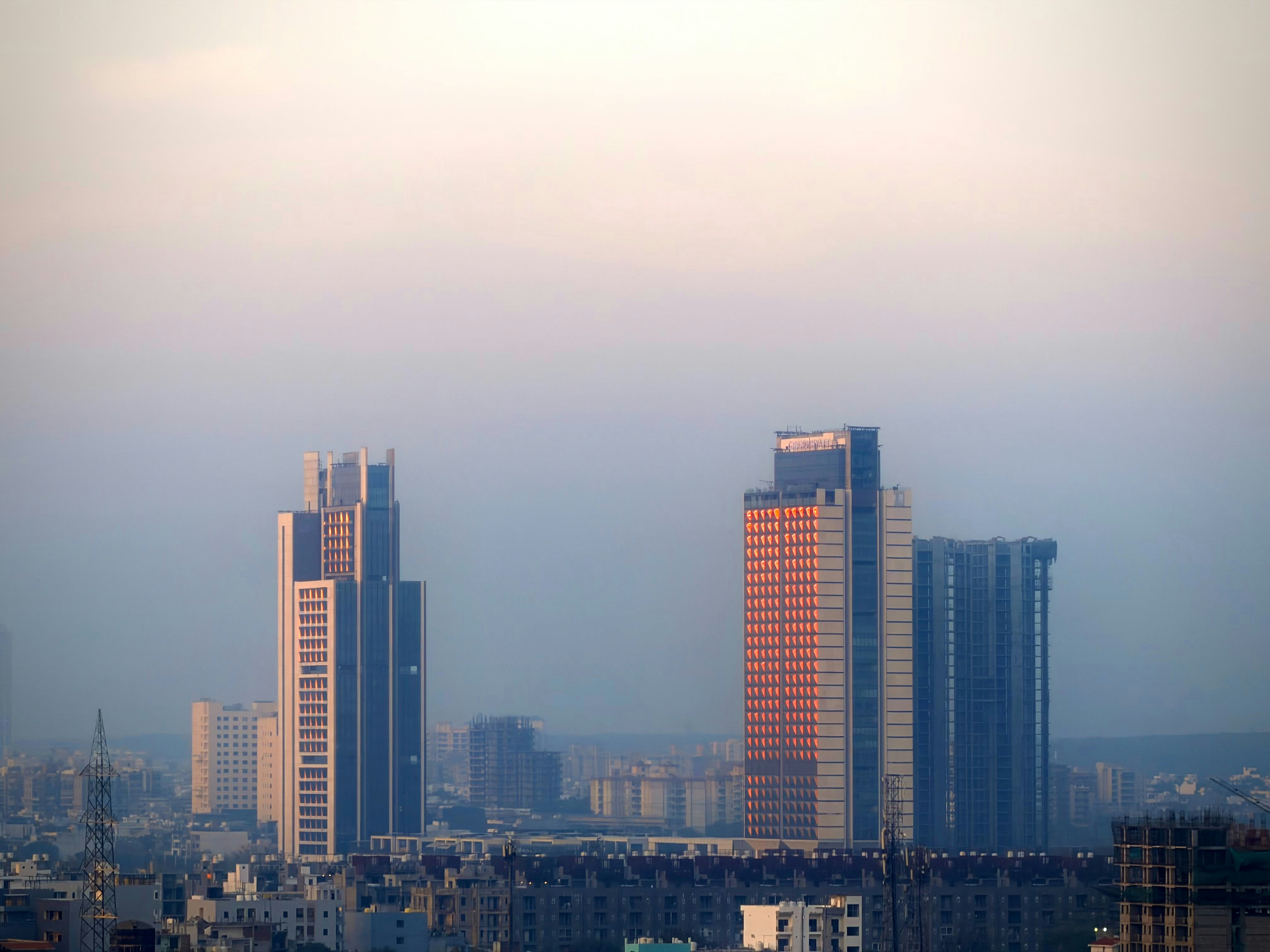 Early-morning cityscape with two prominent high-rises rising above a low-rise skyline; soft pastel sky and distant haze define the scene.