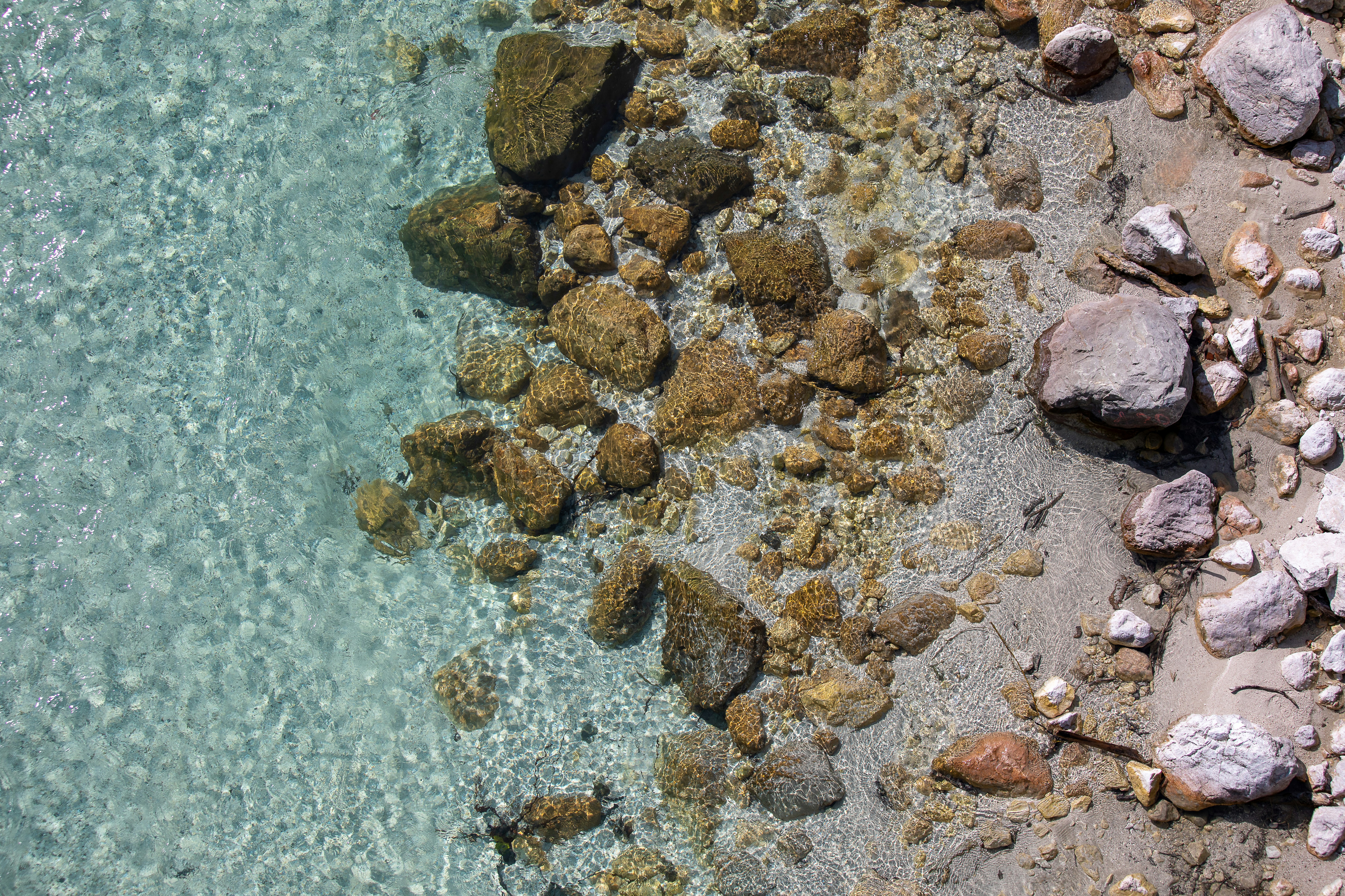 Aerial close-up of the crystal-clear turquoise waters of the Soča River in Slovenia, revealing sun-lit stones and pebbles beneath the surface where the emerald water meets the rocky shoreline.
