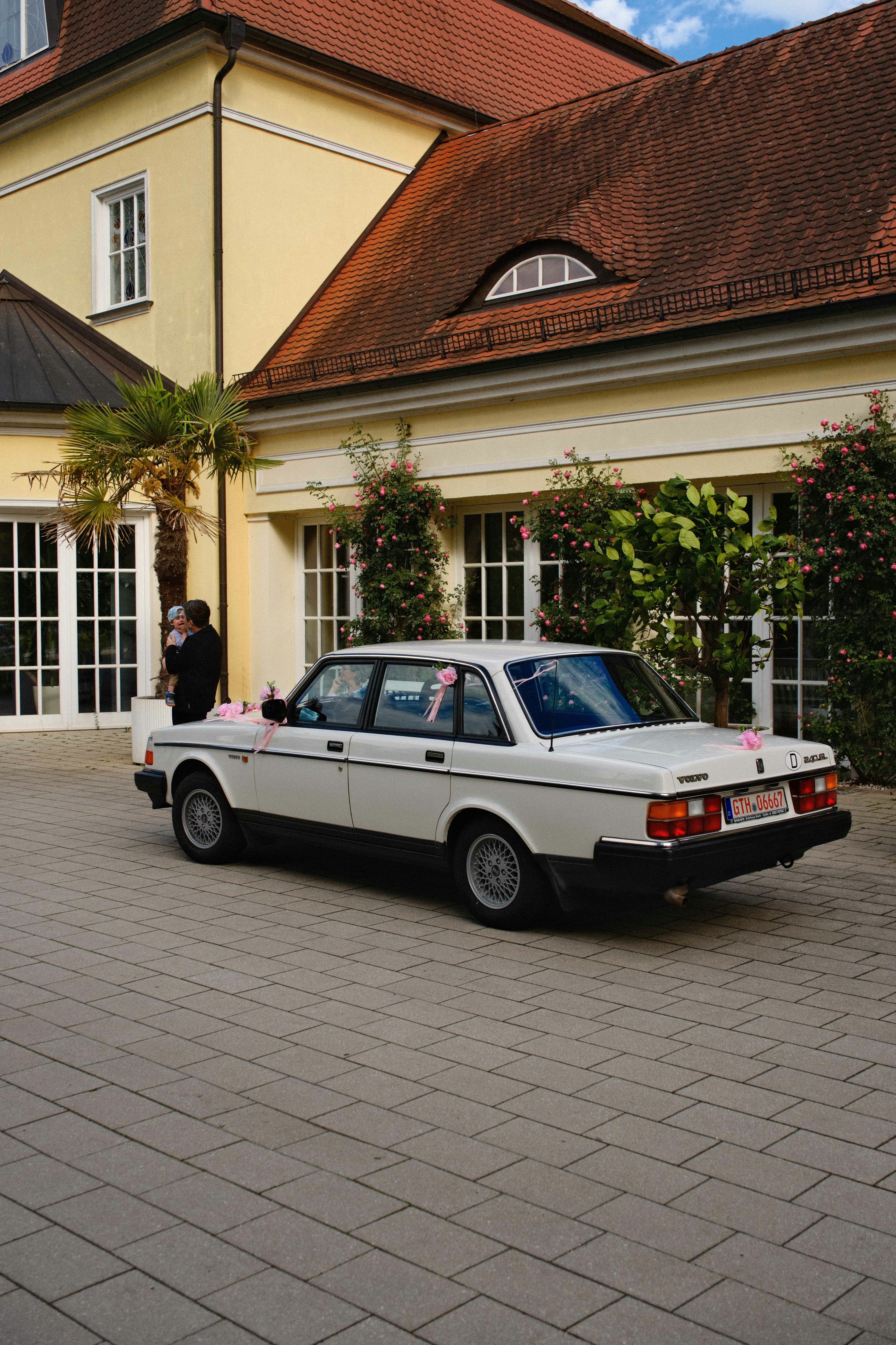 Vintage sedan outside a sunlit villa, with pink ribbons and guests waiting nearby