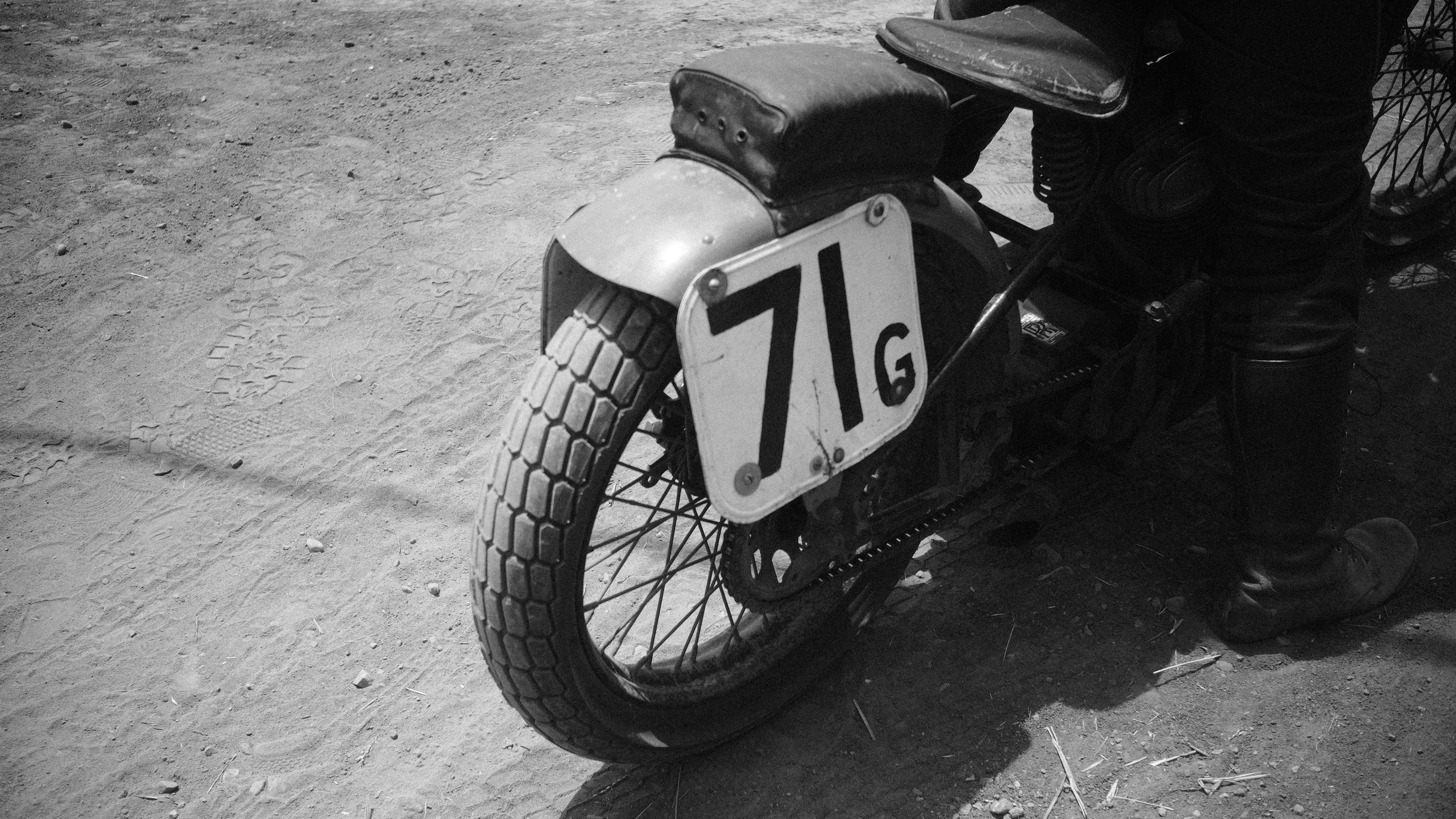 Dark urban motorcycle scene with a close-up on the rear wheel and license plate, captured in black and white to emphasize texture and grit.