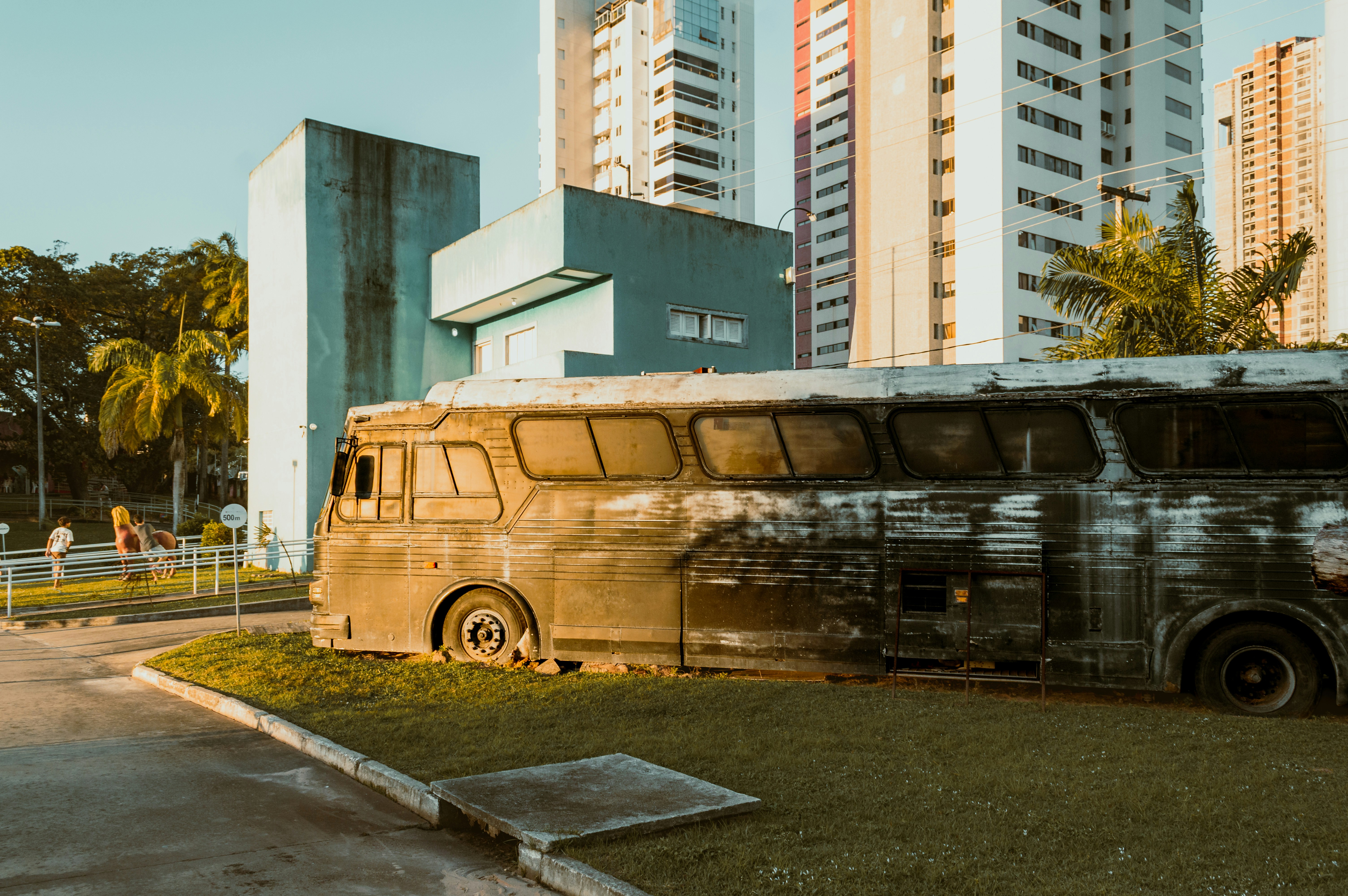 Burnt-out bus parked on grass with tall city buildings.