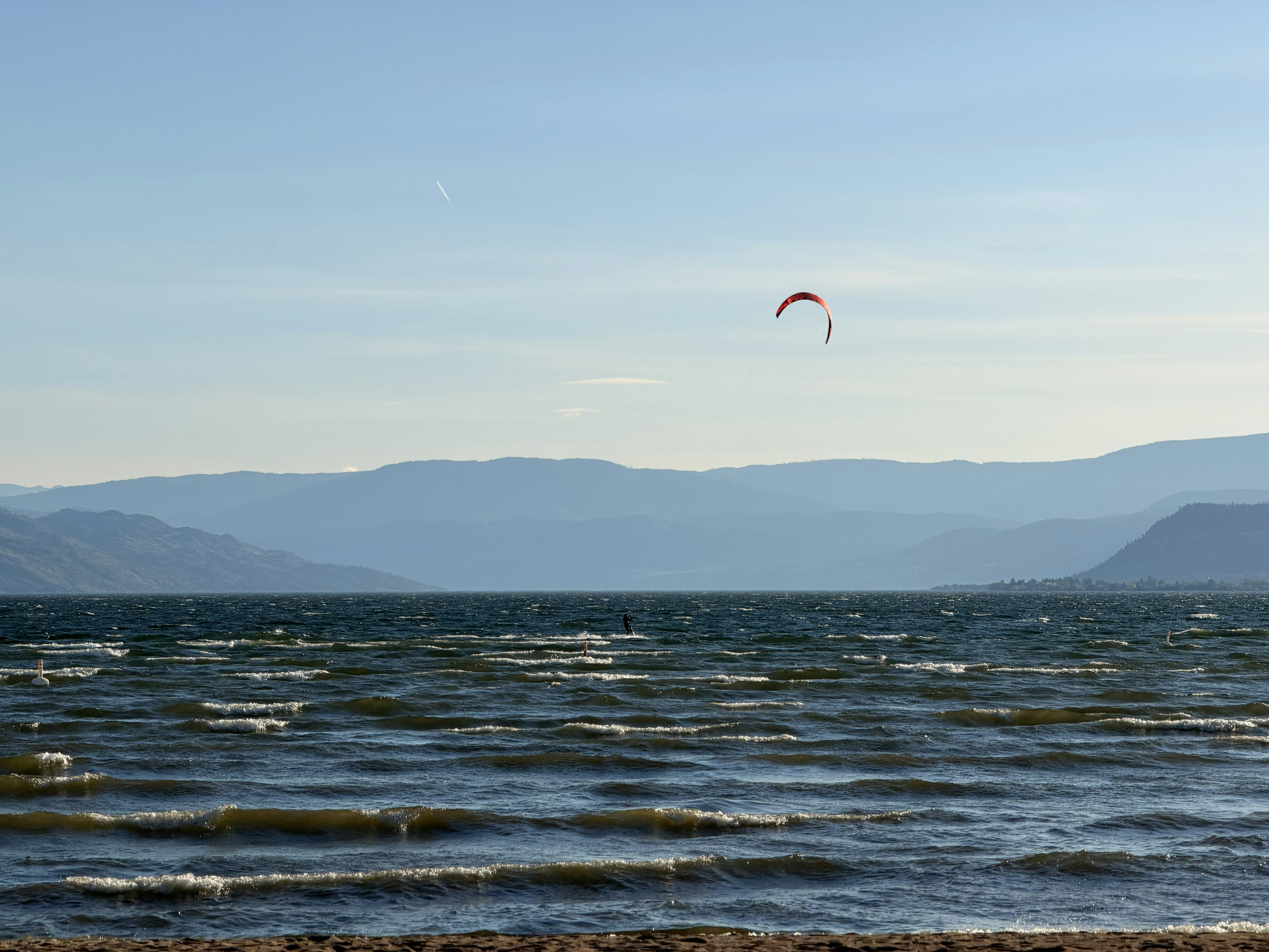 Wide lake with choppy waves and distant mountains under a clear sky; a solitary red paraglider sails above the water, adding a focal point amid the expansive seascape.