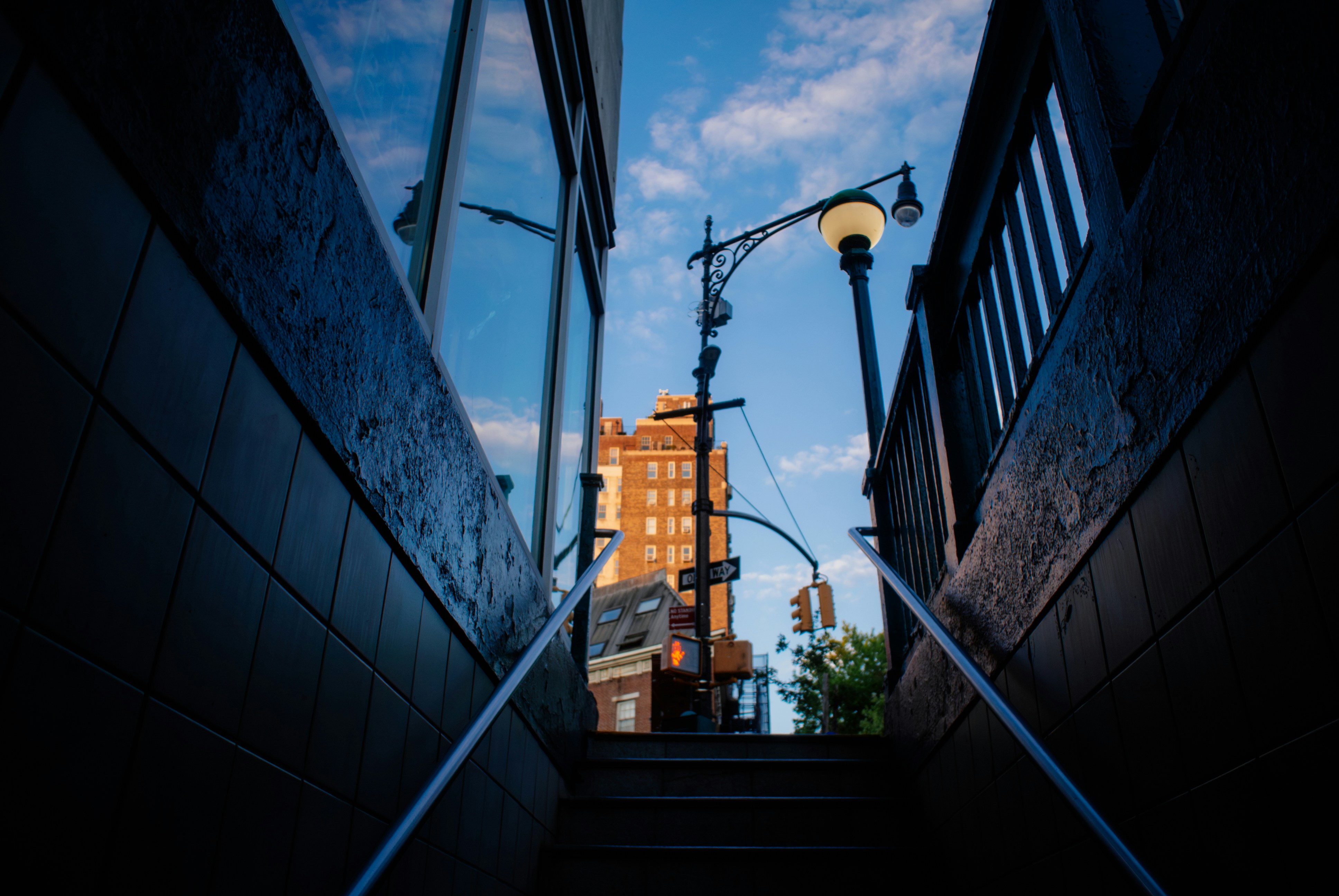A low-angle view up a concrete stairwell toward a city street lamp and buildings, with blue sky and distant tower visible beyond the frame.