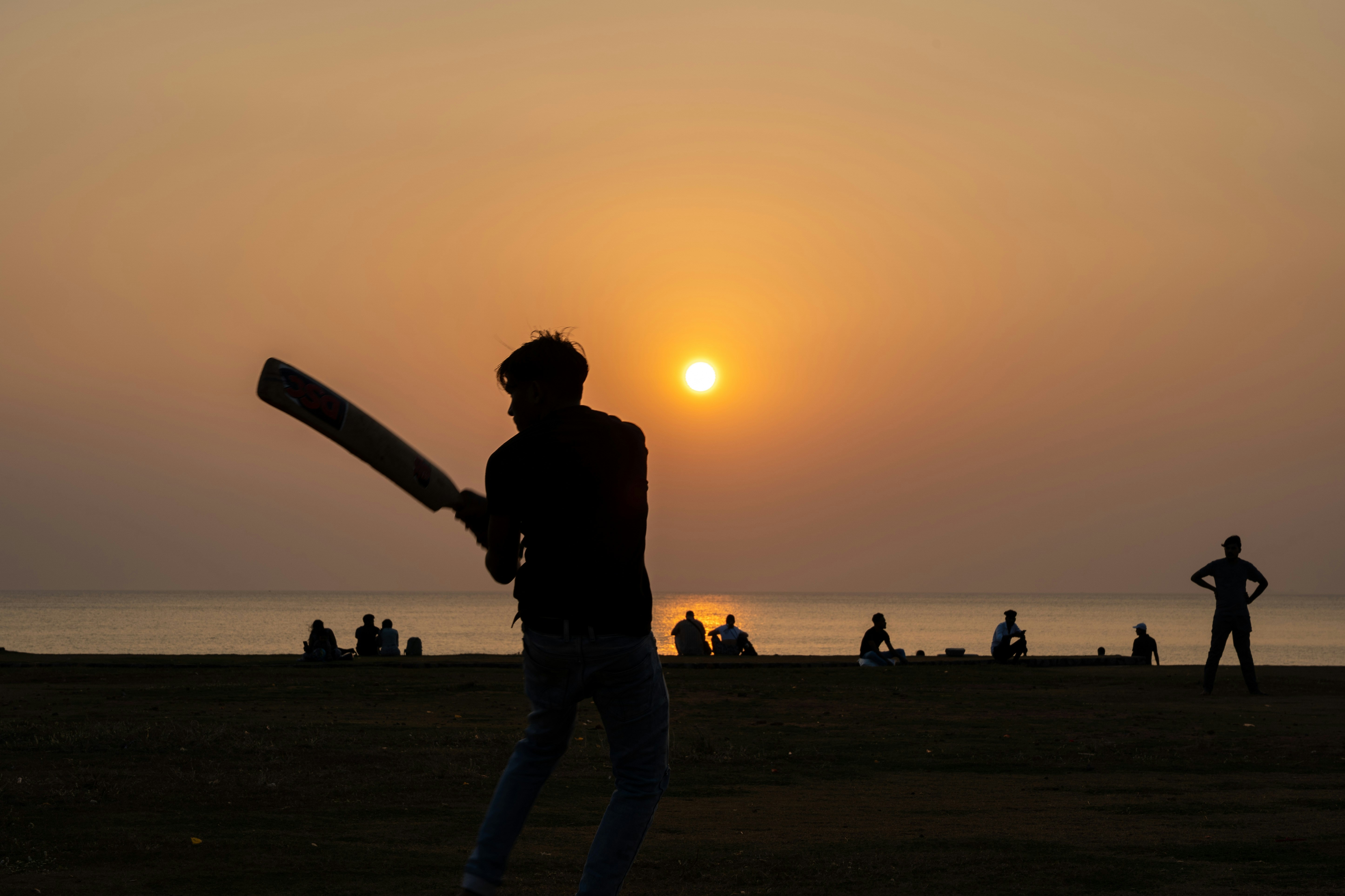 Silhouetted cricket player batting at sunset near the ocean.
