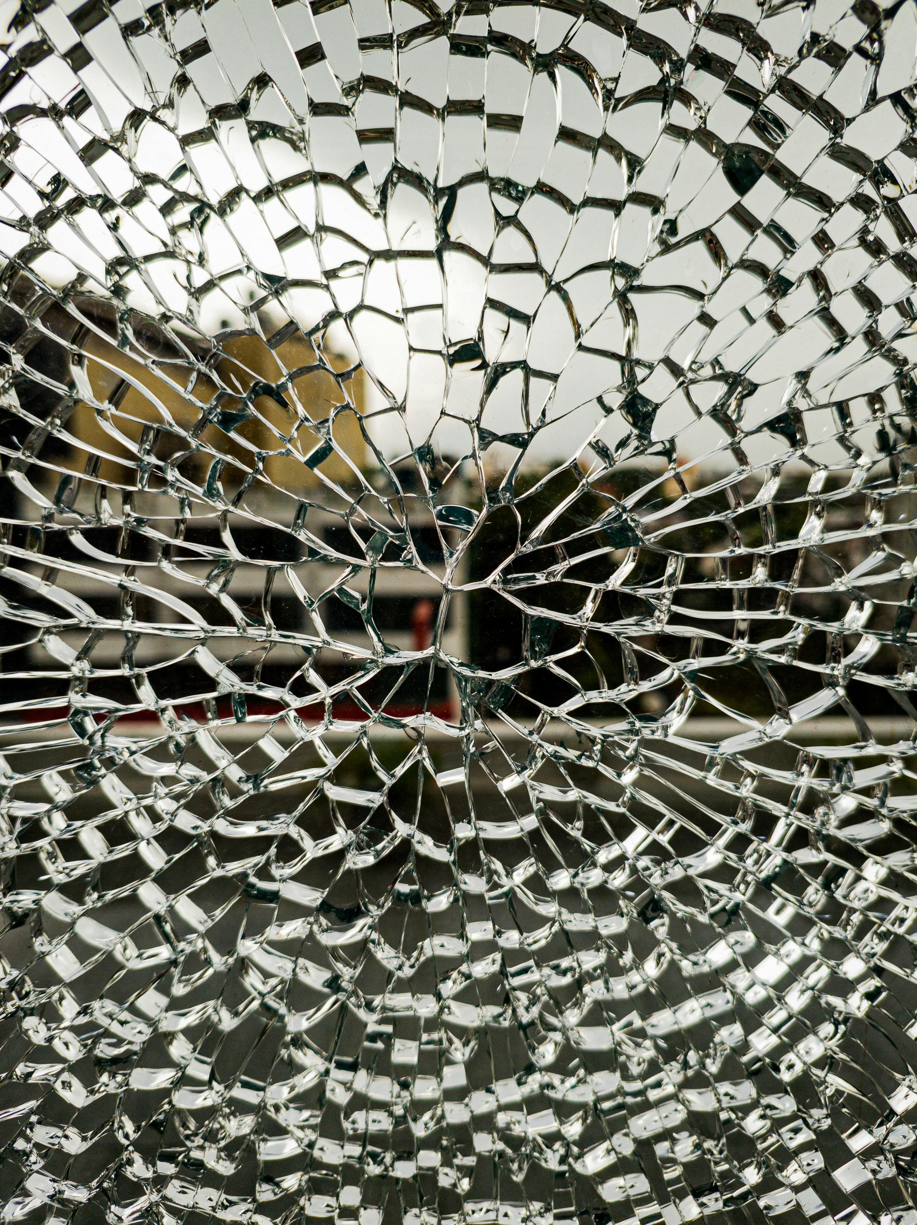 Shattered mirror fills the frame with a central broken-plate pattern, hinting at a distant, blurred background scene. The image emphasizes texture and reflective geometry rather than recognizable detail.
