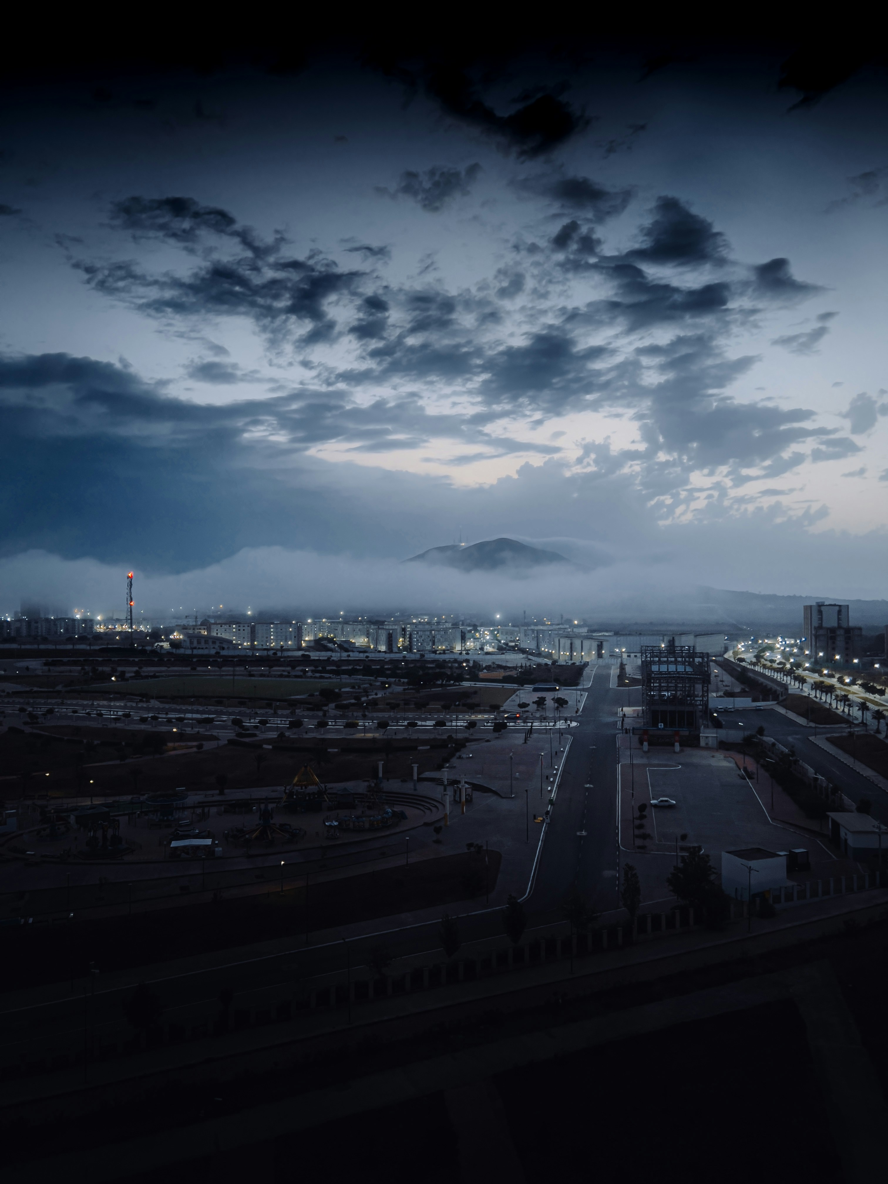 Industrial cityscape at twilight with a distant hill and low-lying fog; the scene emphasizes urban layout and horizon glow rather than close-up subjects.