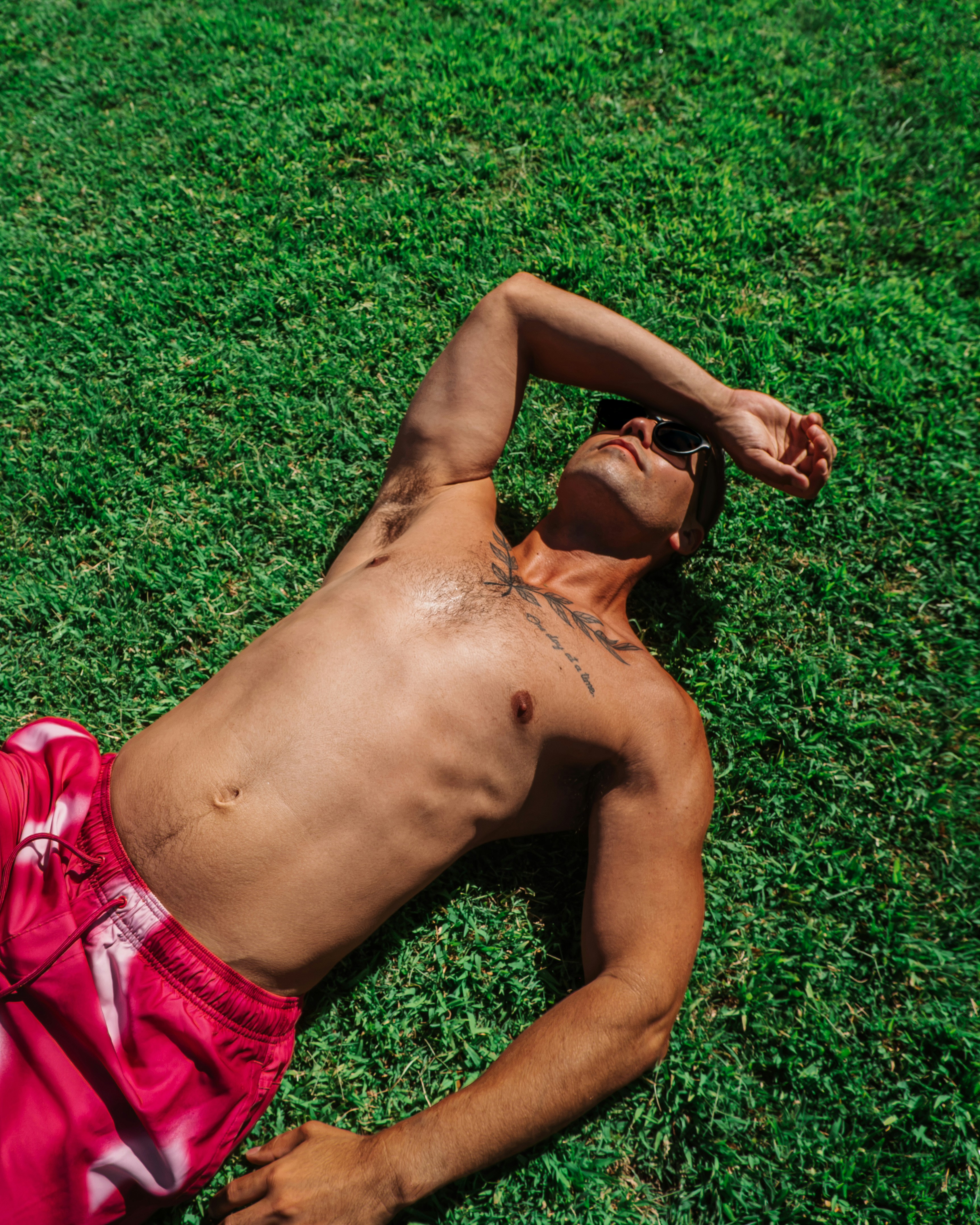 A shirtless man lies on vibrant green grass, wearing pink shorts and sunglasses, basking under sunlight with one arm across his forehead. The scene emphasizes relaxed leisure and sunlit texture.