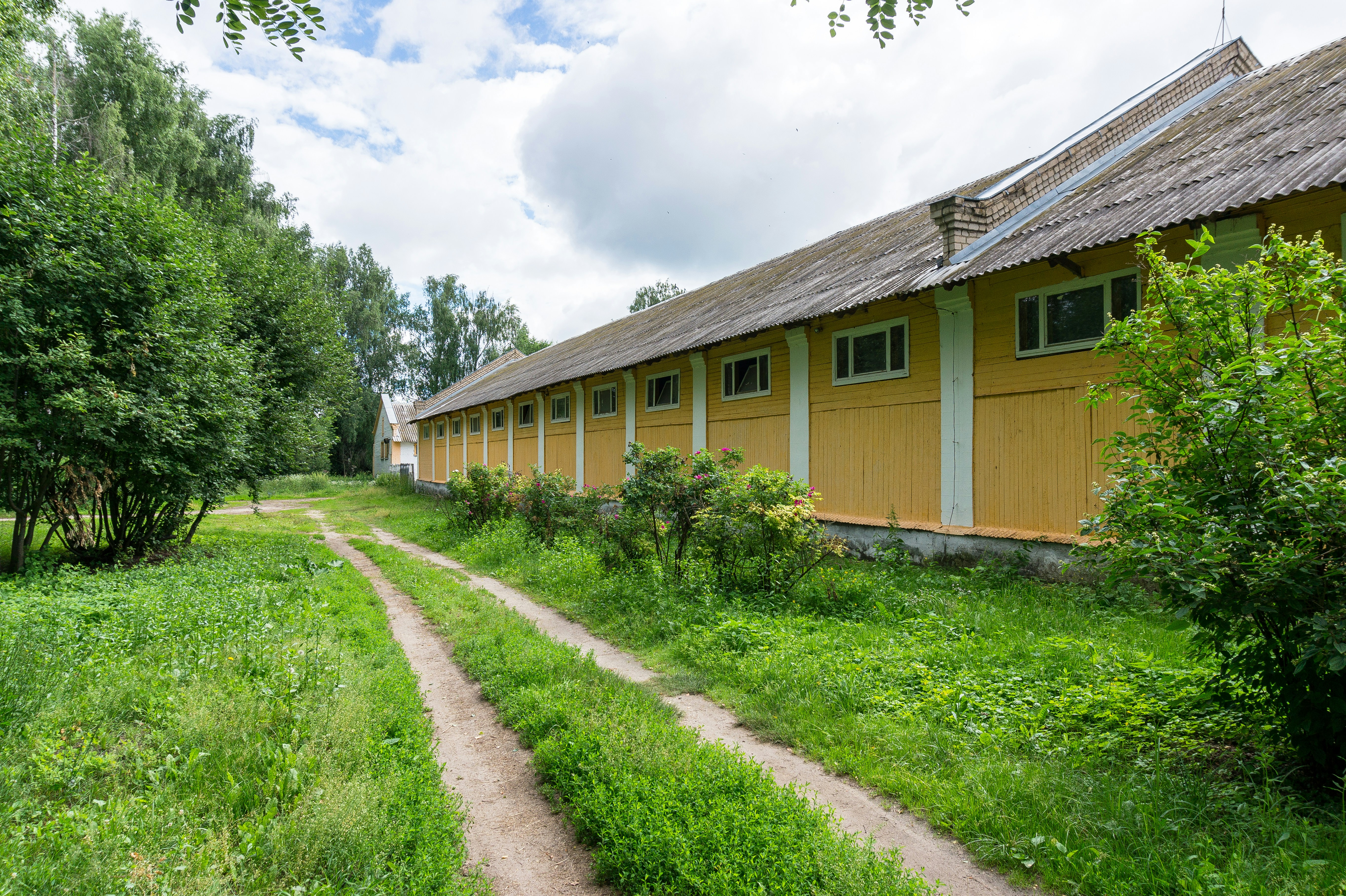 Long wooden farm building surrounded by greenery, with dirt paths leading through the grass