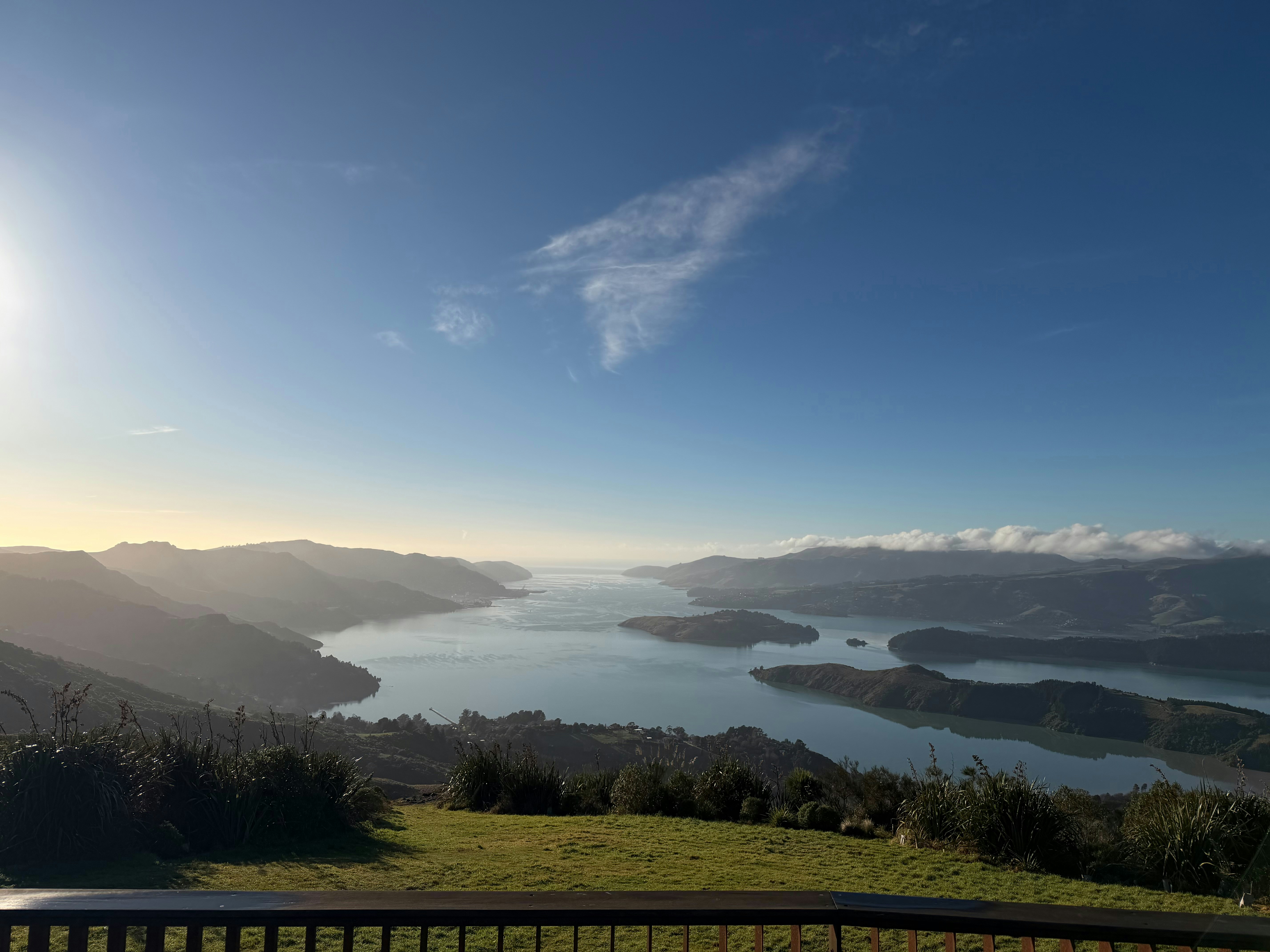Sunlit coastal fjord landscape with layered hills, a calm water body and distant islands; foreground shows a grassy terrace and railing, emphasizing depth and horizon.