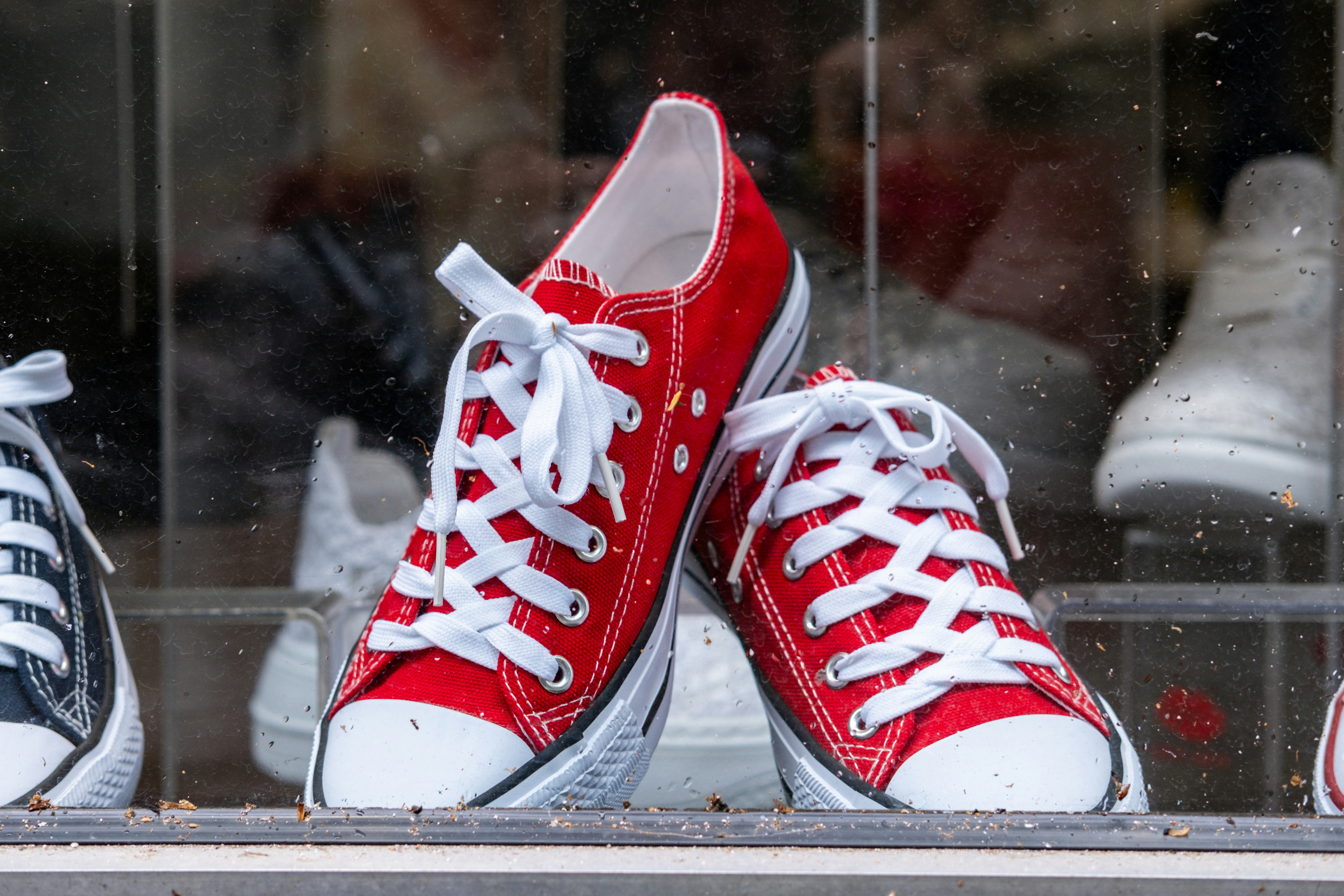 A pair of red canvas sneakers with white laces displayed in a shop window, seen through rain-speckled glass.