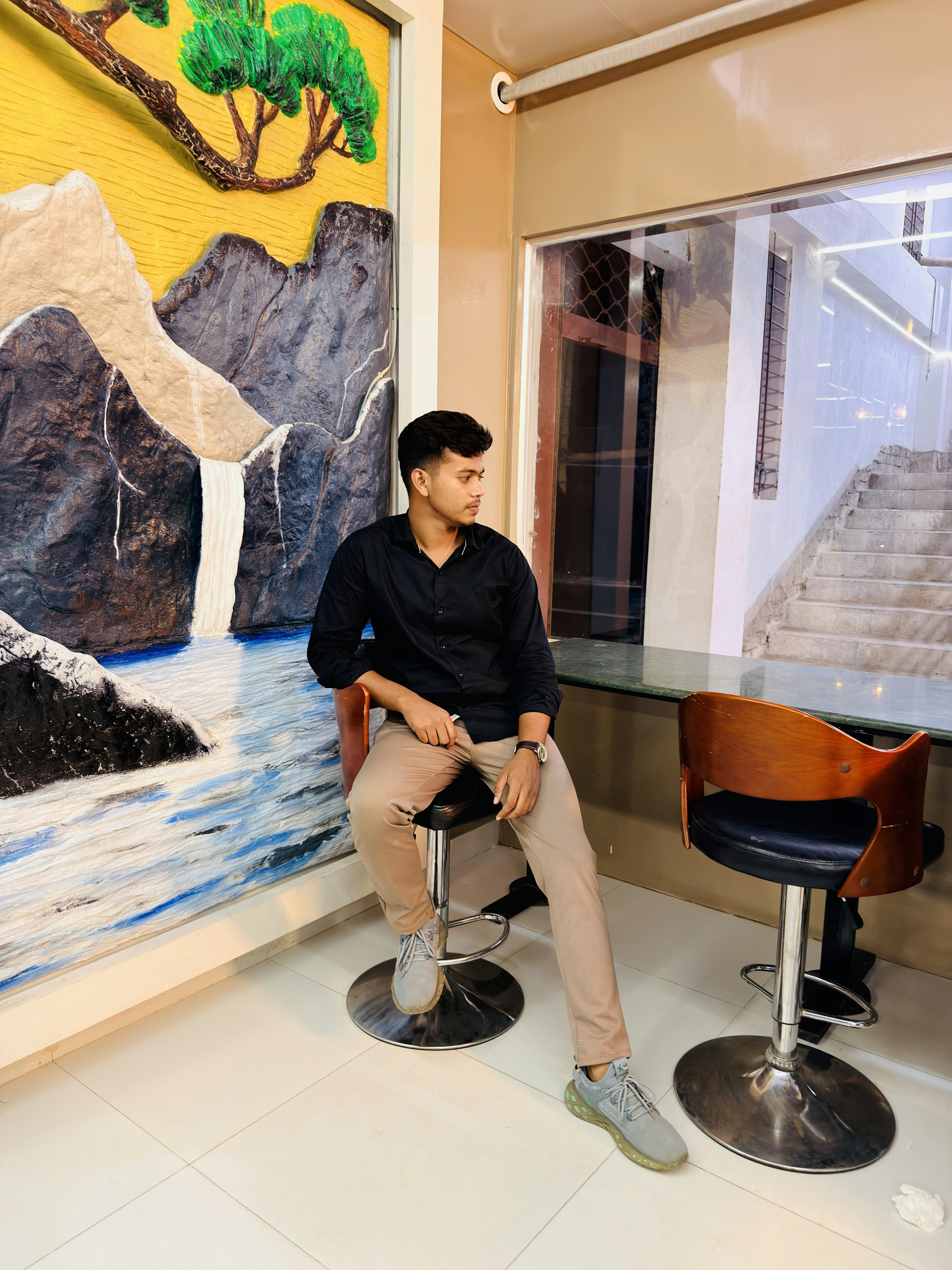 Young man seated at a bar-style counter beside a vivid mural of rocks and a waterfall, with stairs visible through a glass panel in the background.