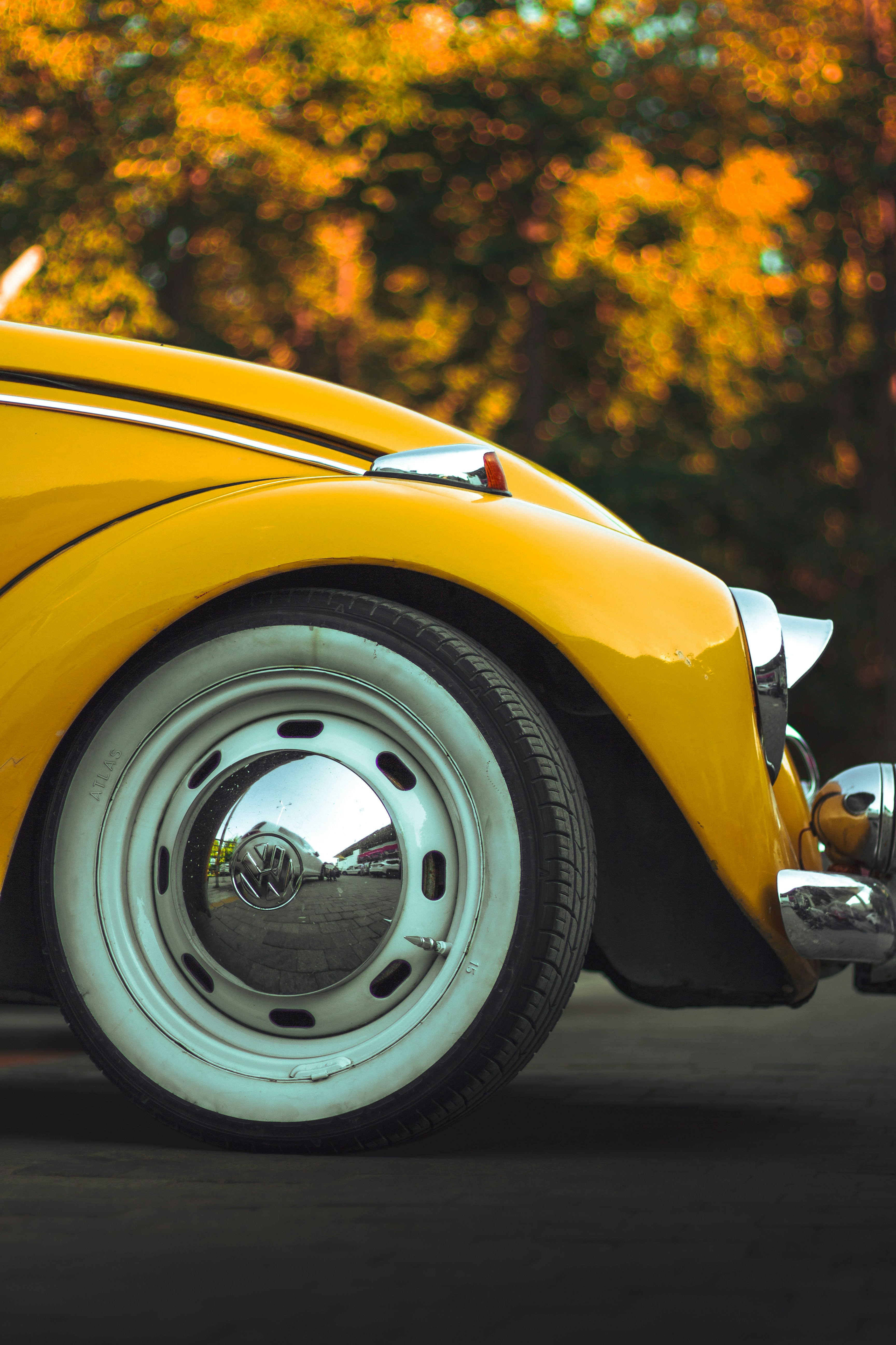 A side-profile cinematic shot of a classic yellow Volkswagen Beetle, focusing on the vintage chrome wheel and whitewall tire with a beautiful background of golden autumn foliage in Bursa, Turkey.