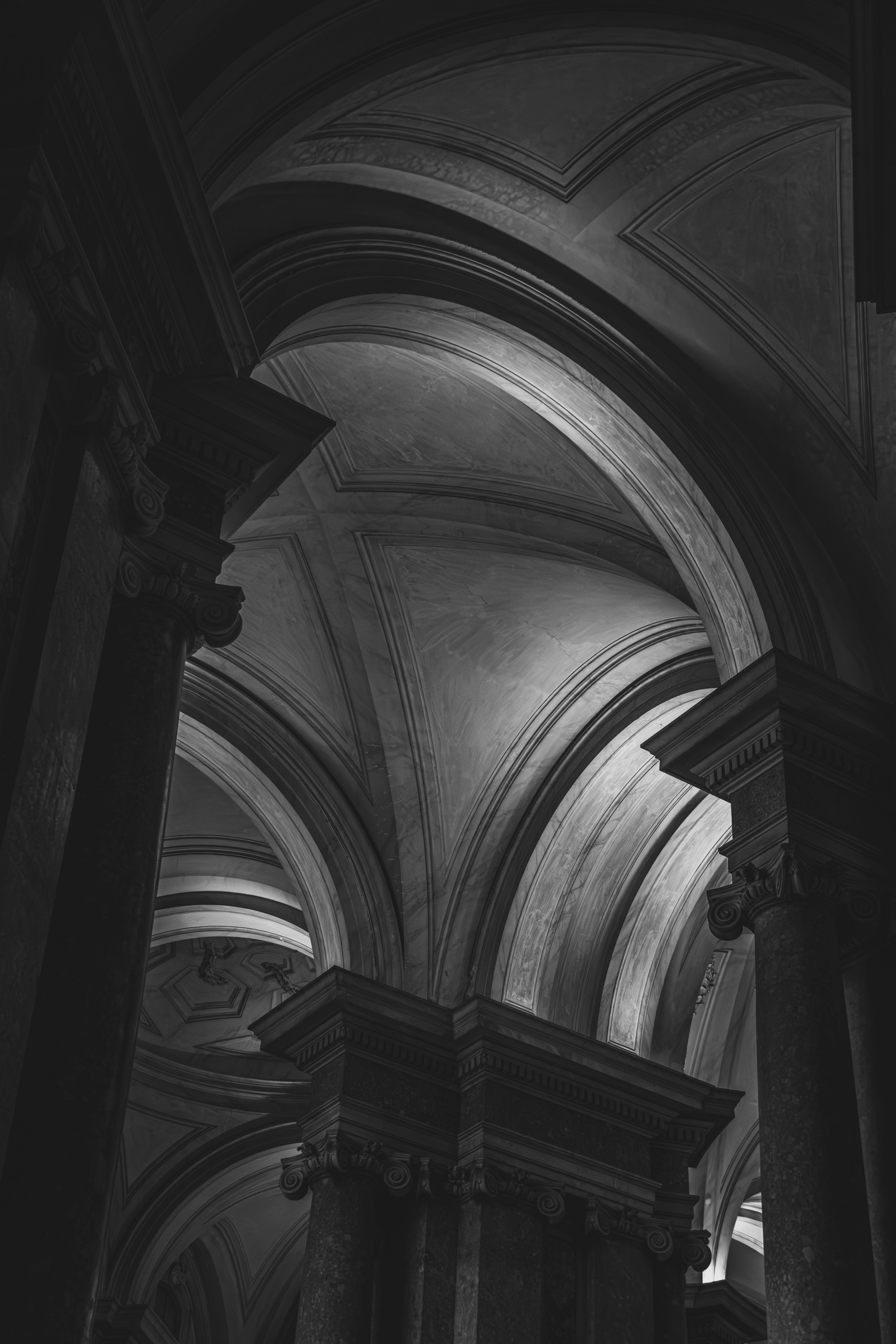 The vaulted marble arches and Ionic columns of the Palace of Caserta in Italy rise into shadow, captured in dramatic black and white. Light falls across the barrel vaults while the columns recede into darkness, giving the grand interior a moody, cathedral-like weight.