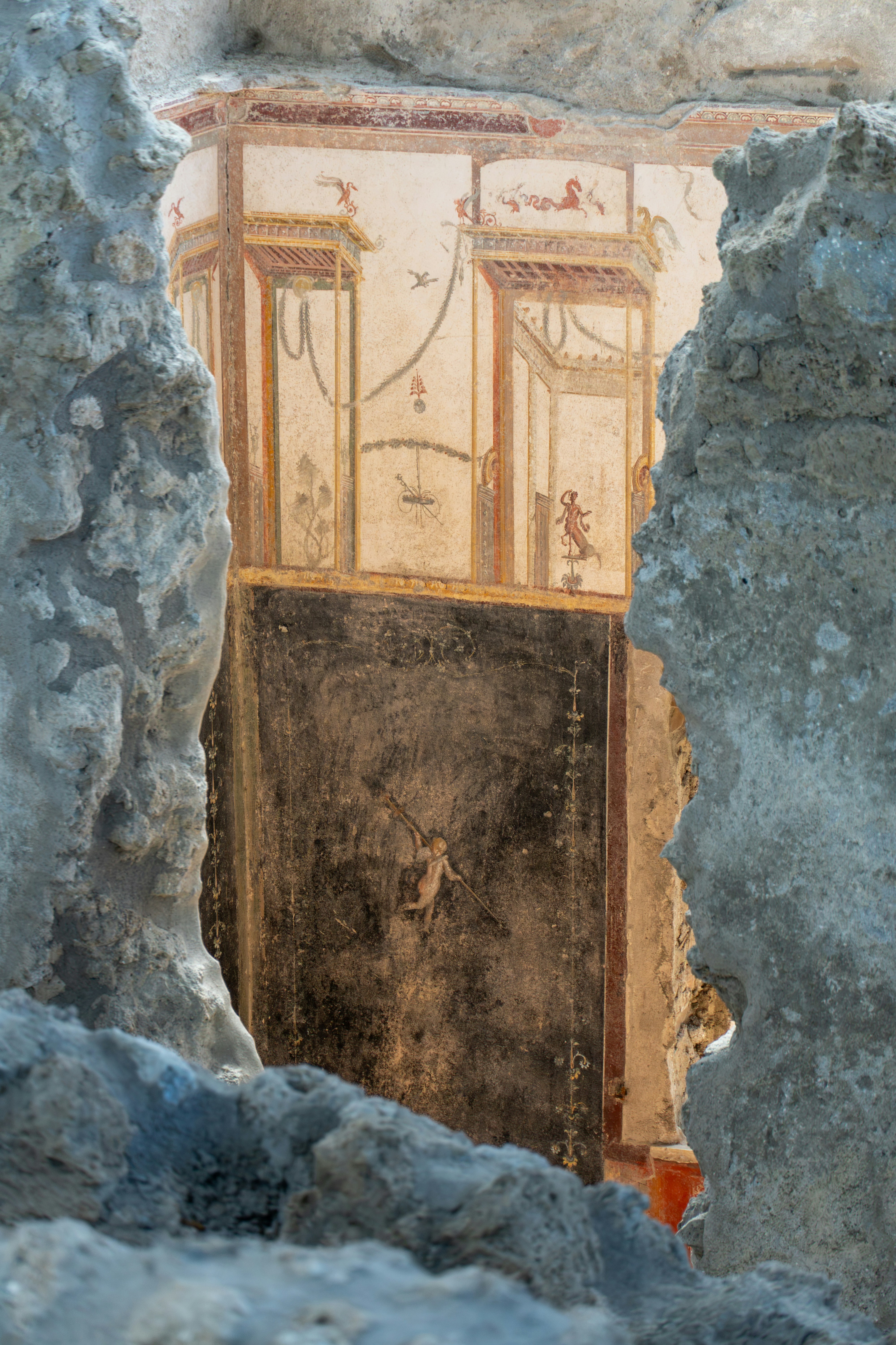 A delicate Roman fresco glimpsed through a rough gap in a stone wall at Pompeii, Italy, its fine painted architecture, garlands and mythological figures still vivid after nearly two thousand years. The jagged opening frames the ancient artwork like a natural window into the buried city.