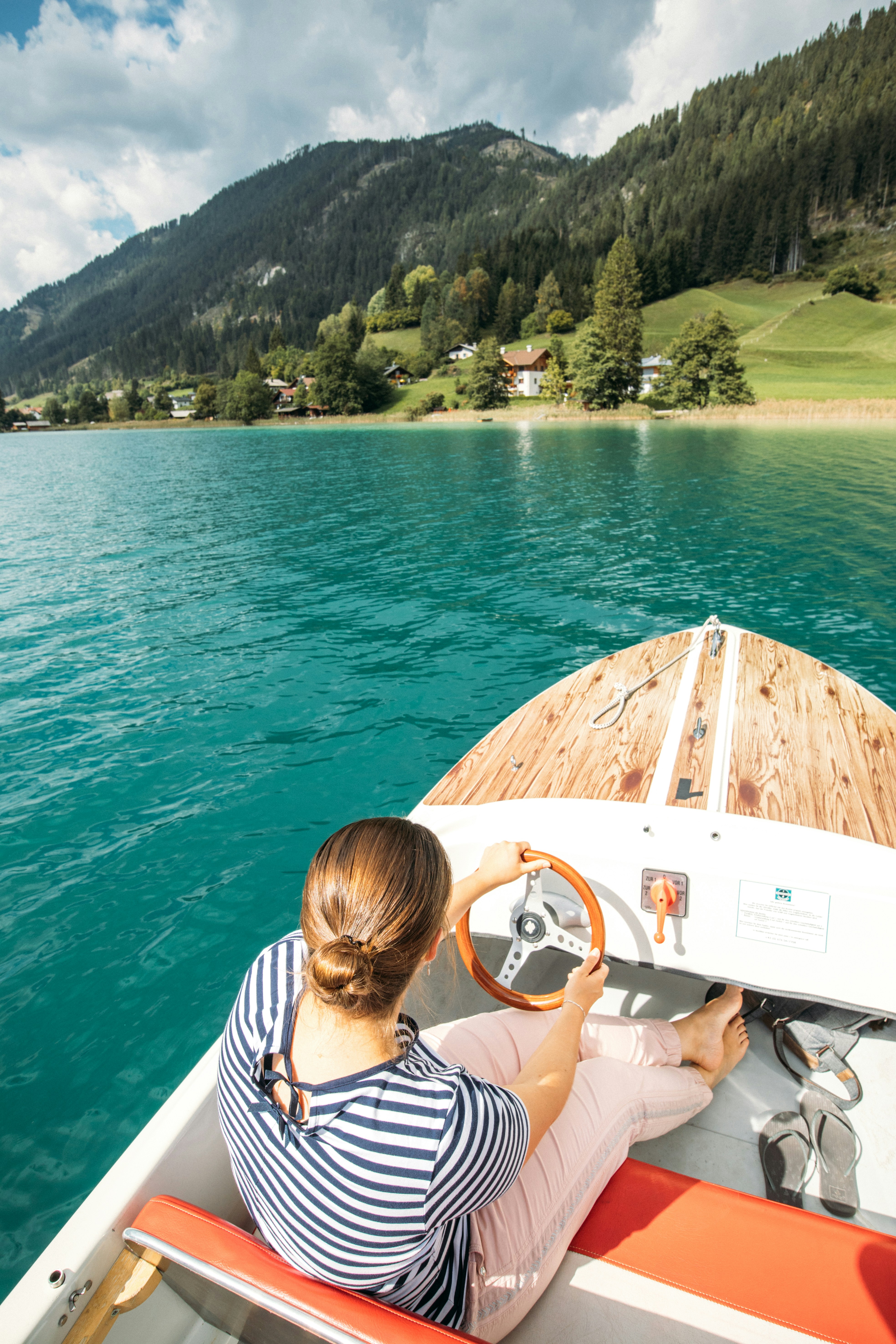 A woman steers a small wooden boat across a turquoise lake with a lakeside village and forested hills in the distance.