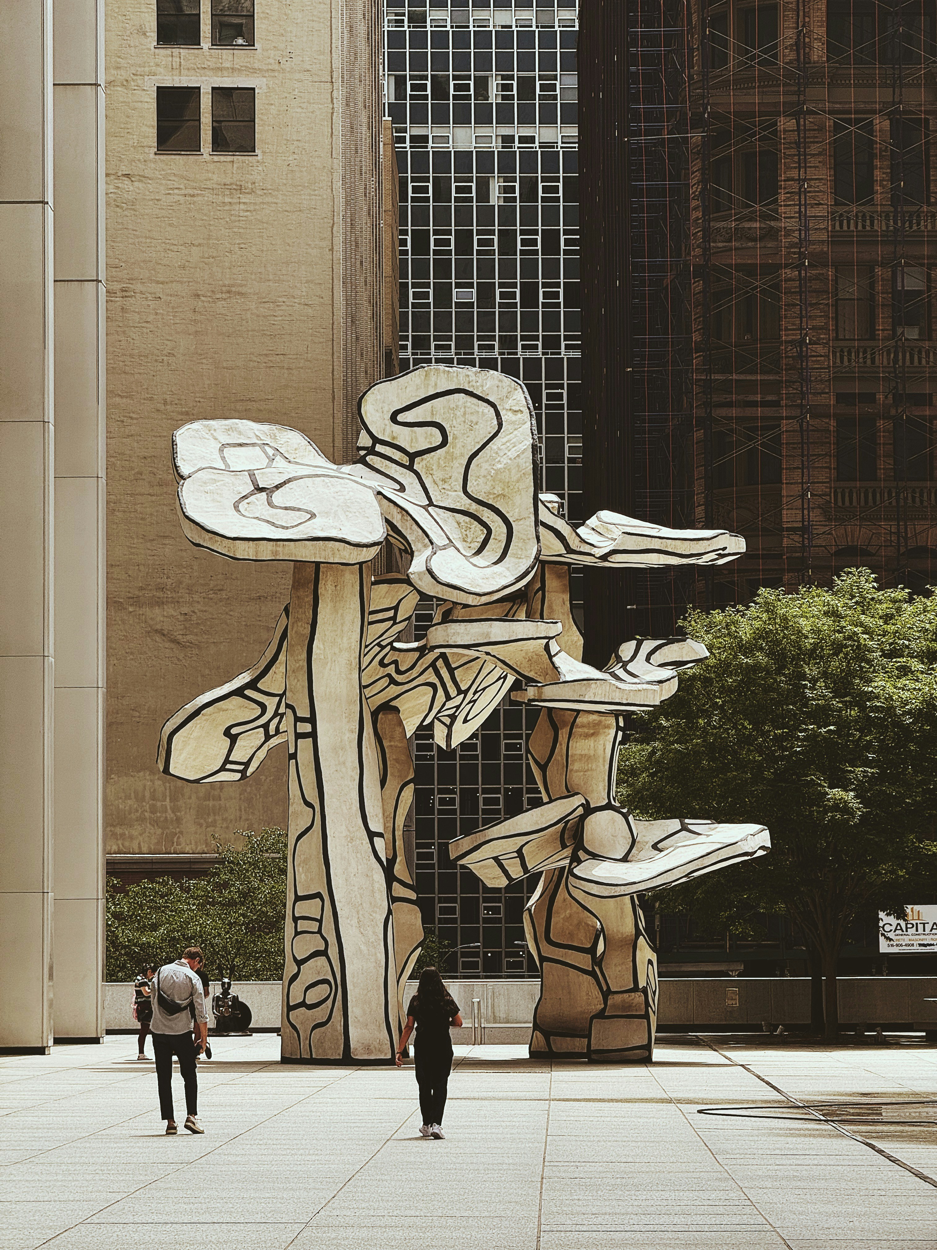 Abstract steel sculpture stands in an urban plaza with two pedestrians nearby; modern buildings frame the scene and a tree adds color contrast. Public art installation captured in daylight.