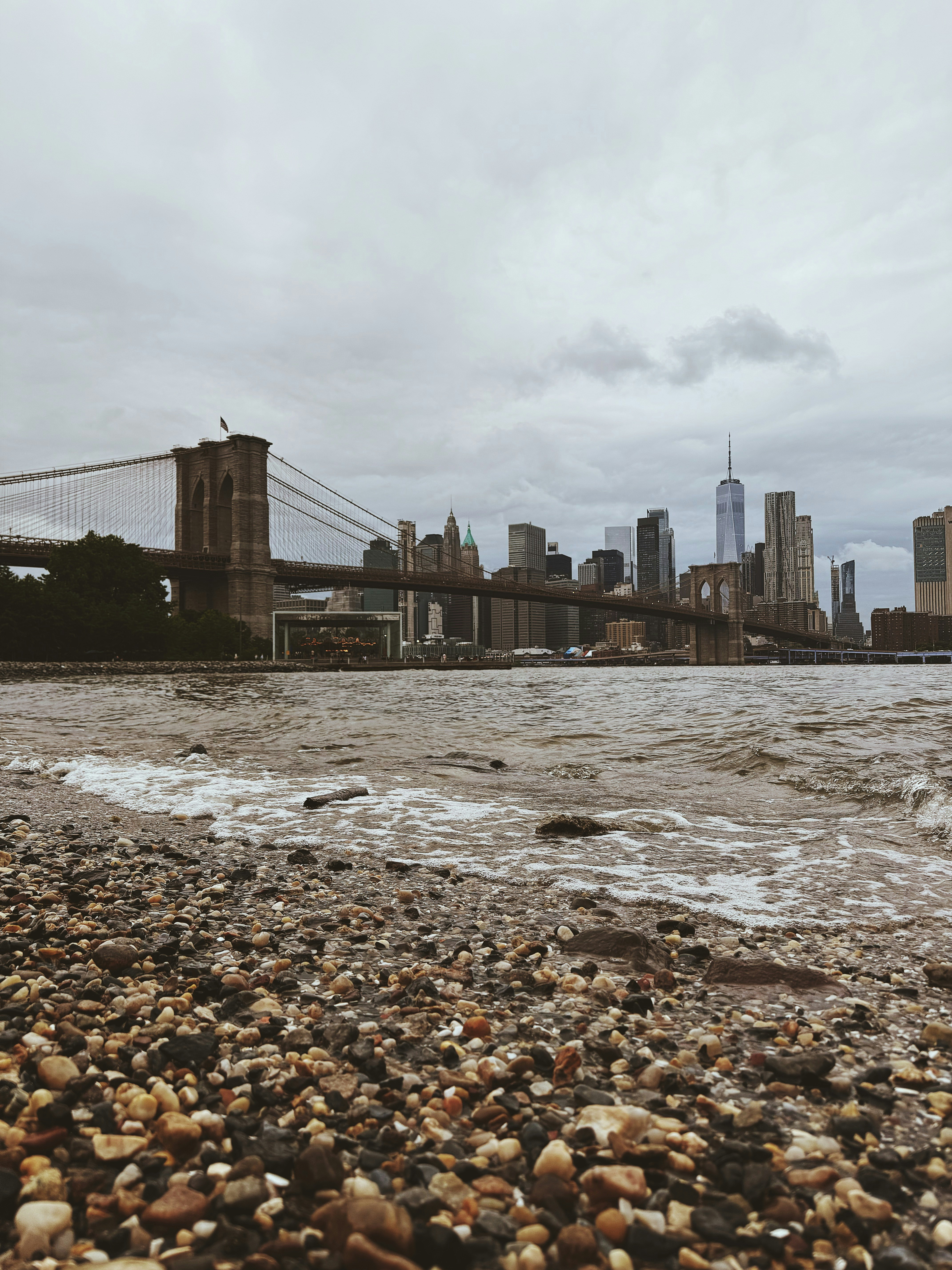 Brooklyn Bridge, Lower Manhattan, and pebbled riverbank under a dramatic overcast sky