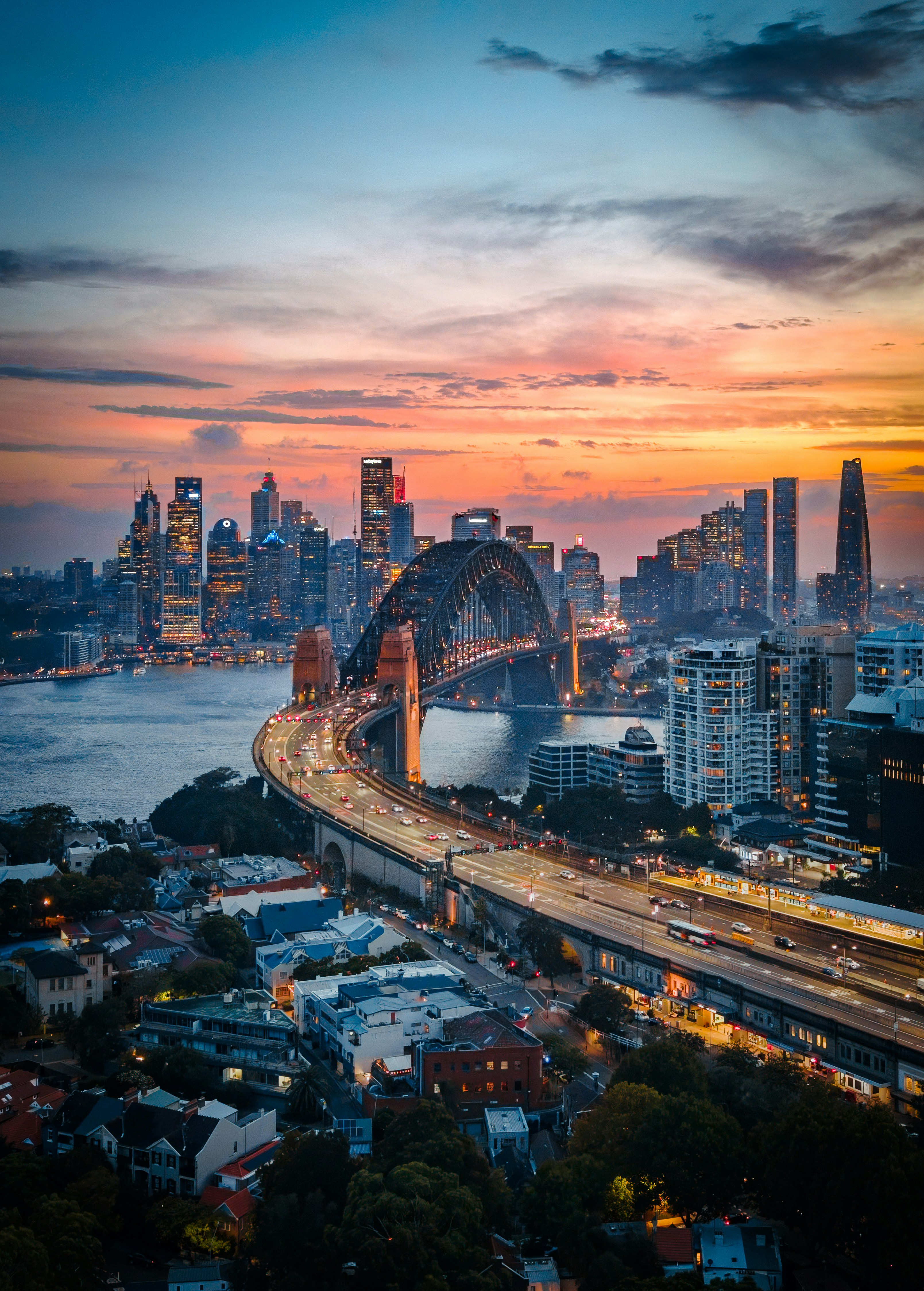 City sunset over a curving harbor bridge with illuminated skyline and road traffic; urban landscape showing high-rise buildings and residential blocks at dusk. (Photograph) 