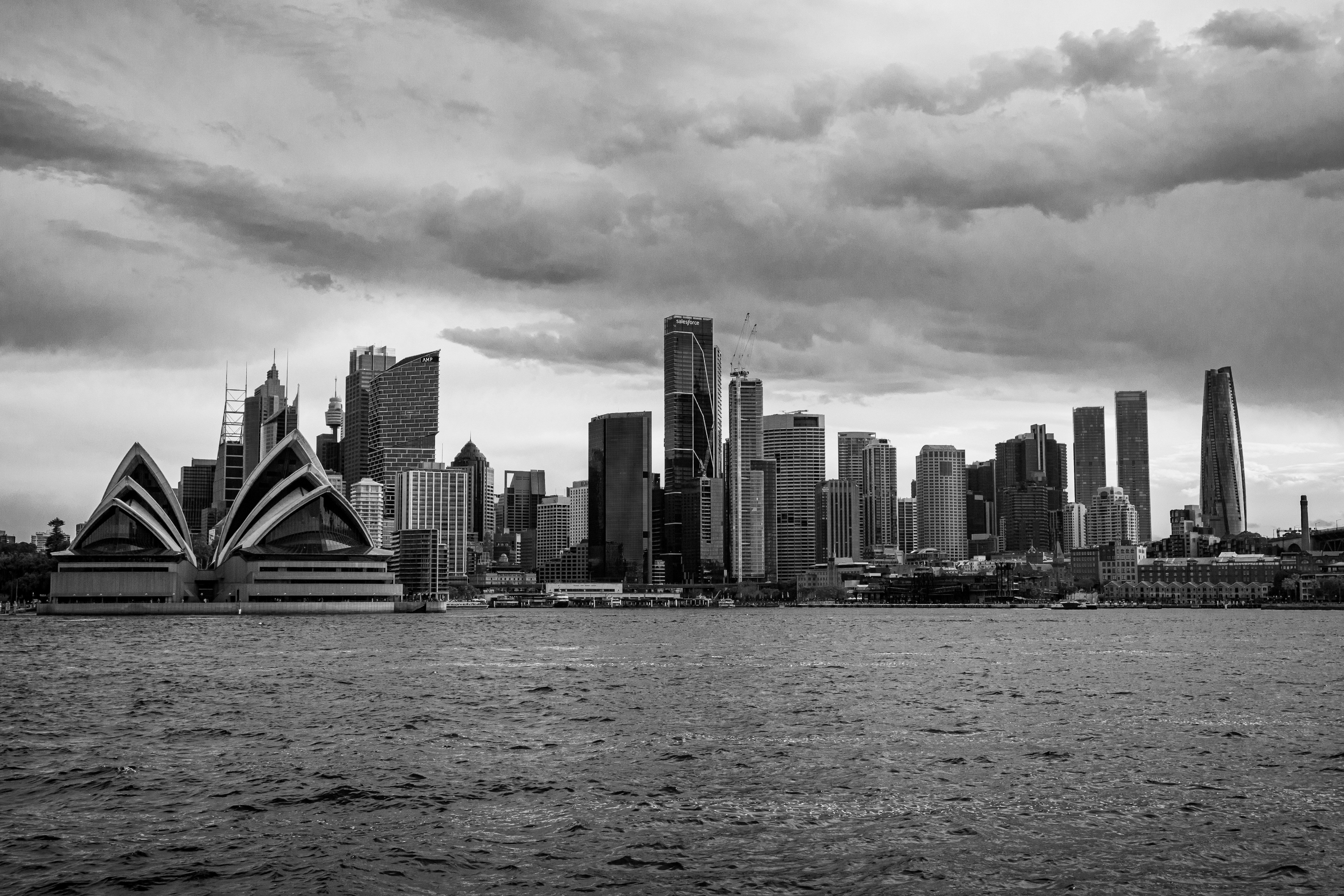 Monochrome view of the Sydney skyline across the harbour with the Opera House on the left and tall buildings rising behind; water in the foreground and dramatic clouds above.