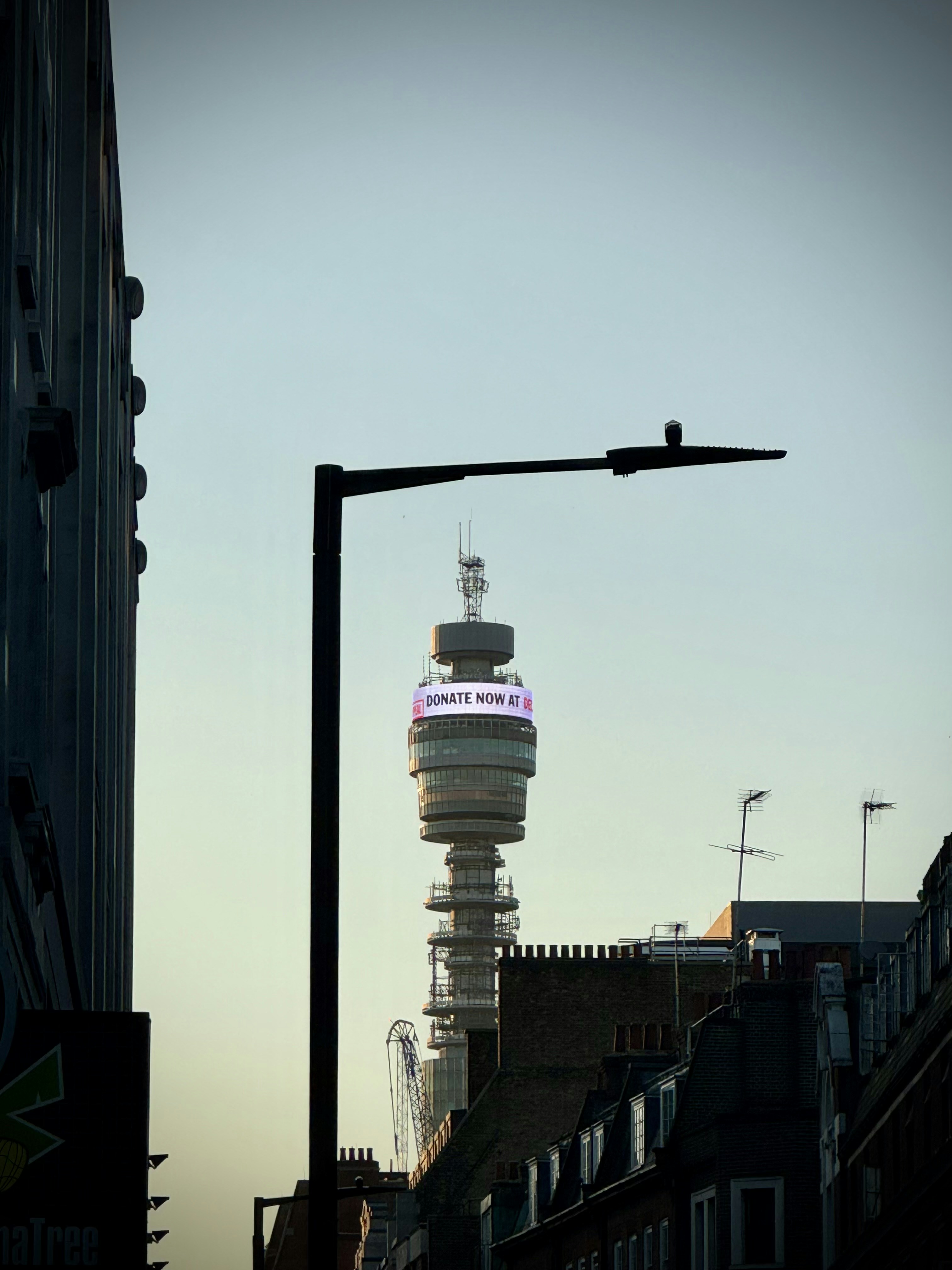 Urban dusk scene with a prominent telecommunications tower rising between dark building silhouettes; a street lamp foreground frames the composition.