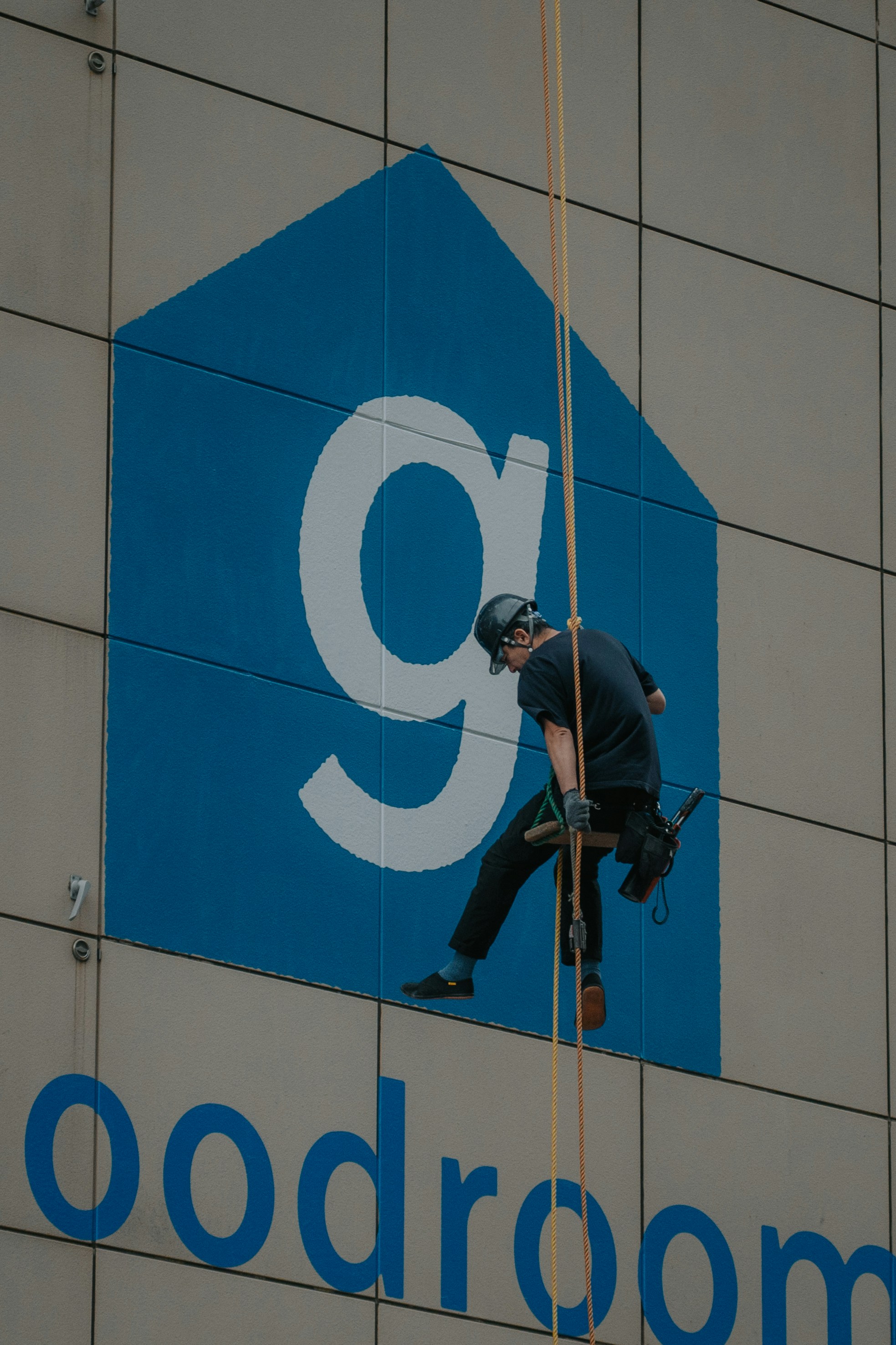 Climber suspended on a rope ascends a tall exterior wall with a large blue sign and oversized number; tools at the belt indicate active construction/maintenance work.