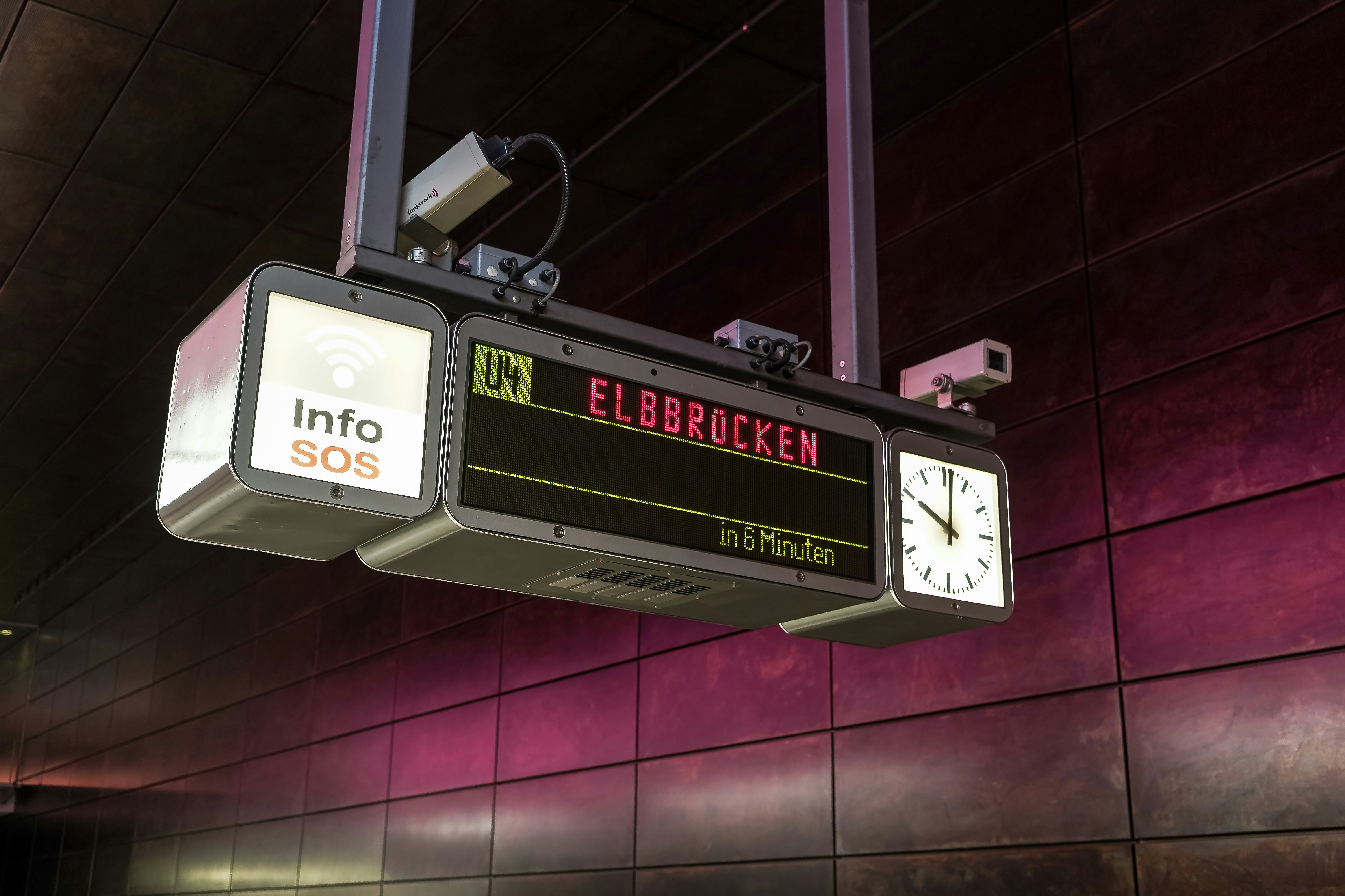 Overhead digital sign and clock at a dimly lit subway station with a magenta-tinted wall, capturing a moment of transit ambience.